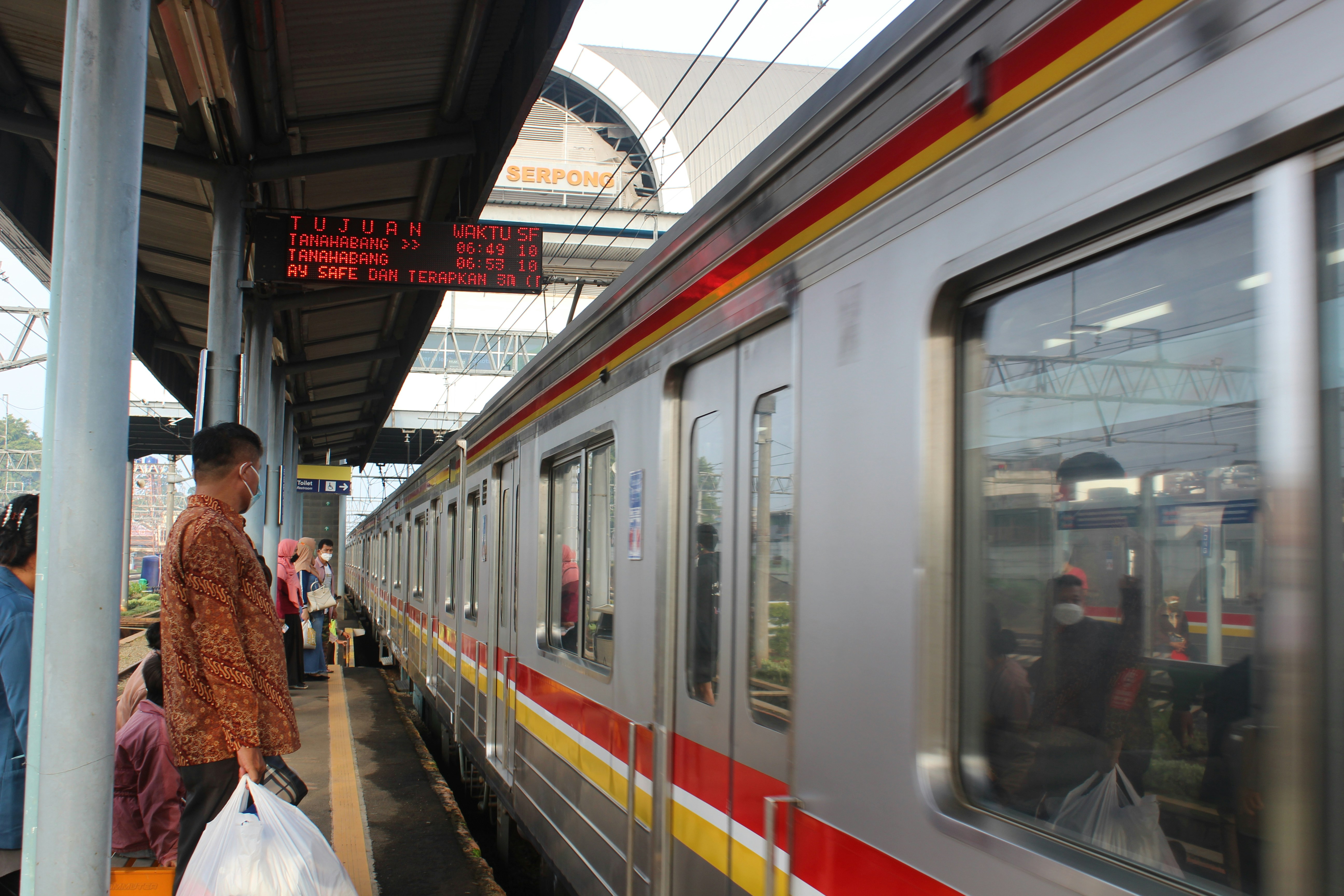 A busy train station scene featuring a commuter train and passengers waiting on the platform. The electronic display shows train destinations and times.