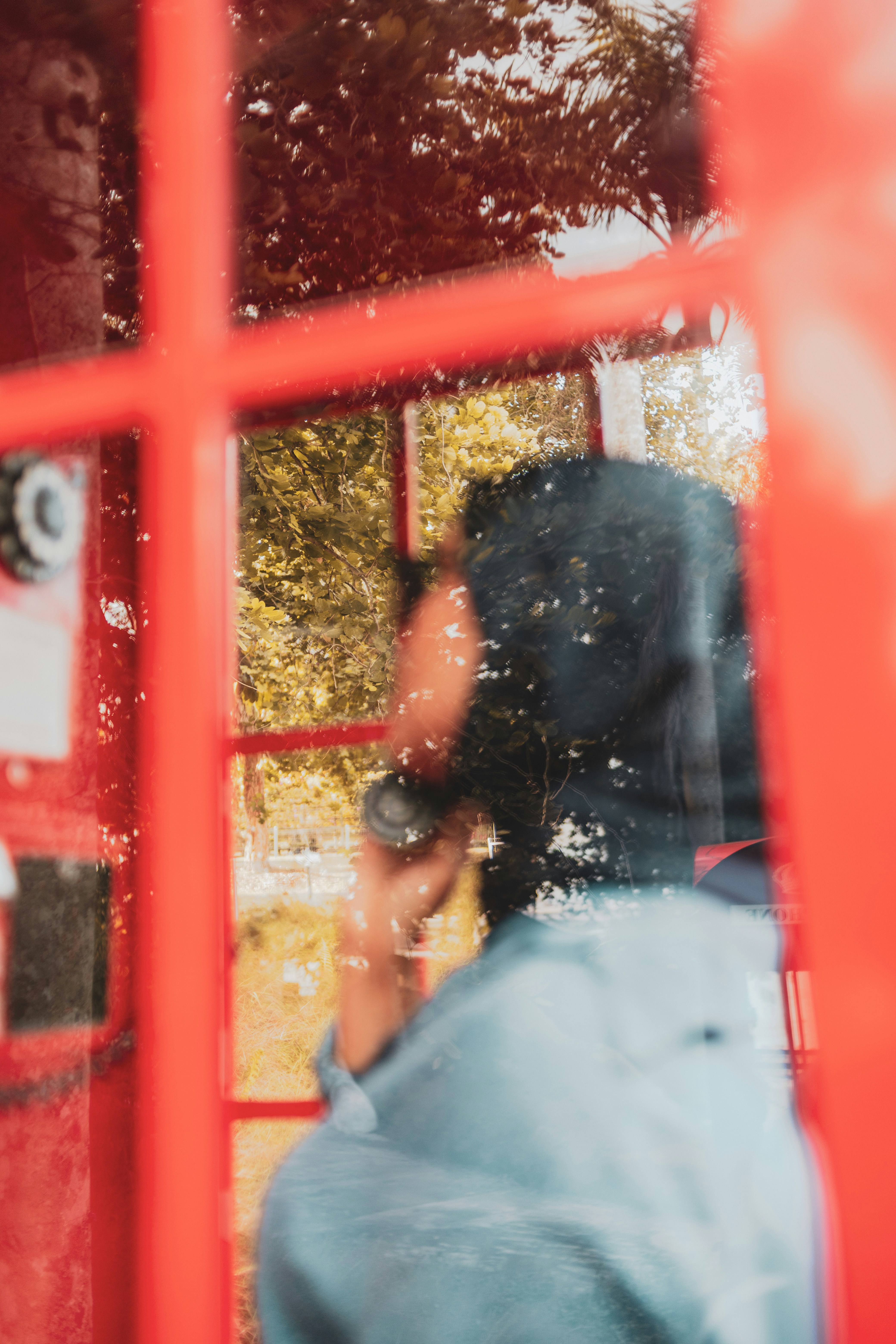 A person engaged in a phone call inside a vintage red telephone booth, with reflections of autumn foliage creating a dreamy atmosphere.