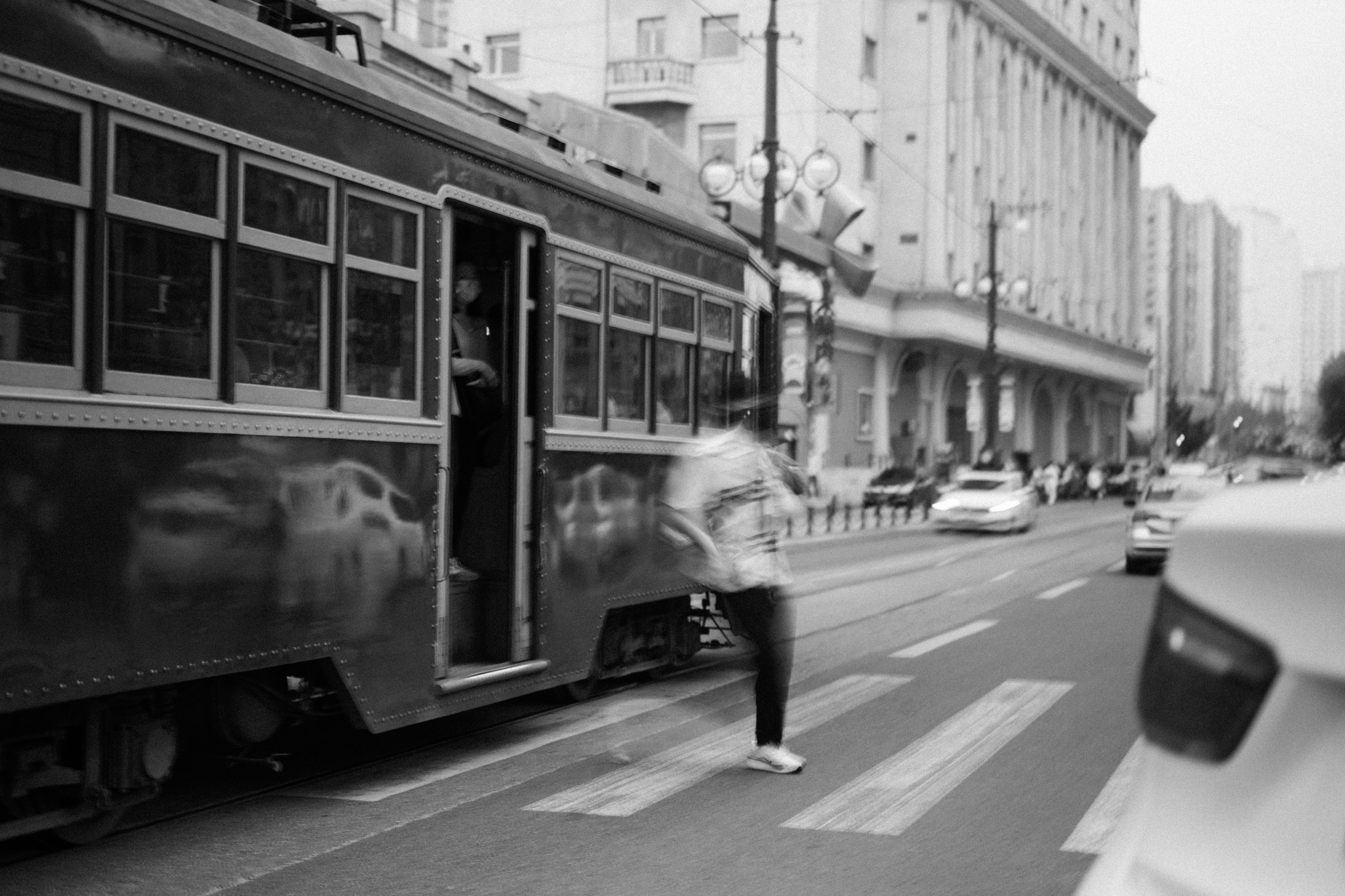 A vintage tram pauses at a bustling street corner as a pedestrian strides across the crosswalk, capturing the essence of urban life in motion.