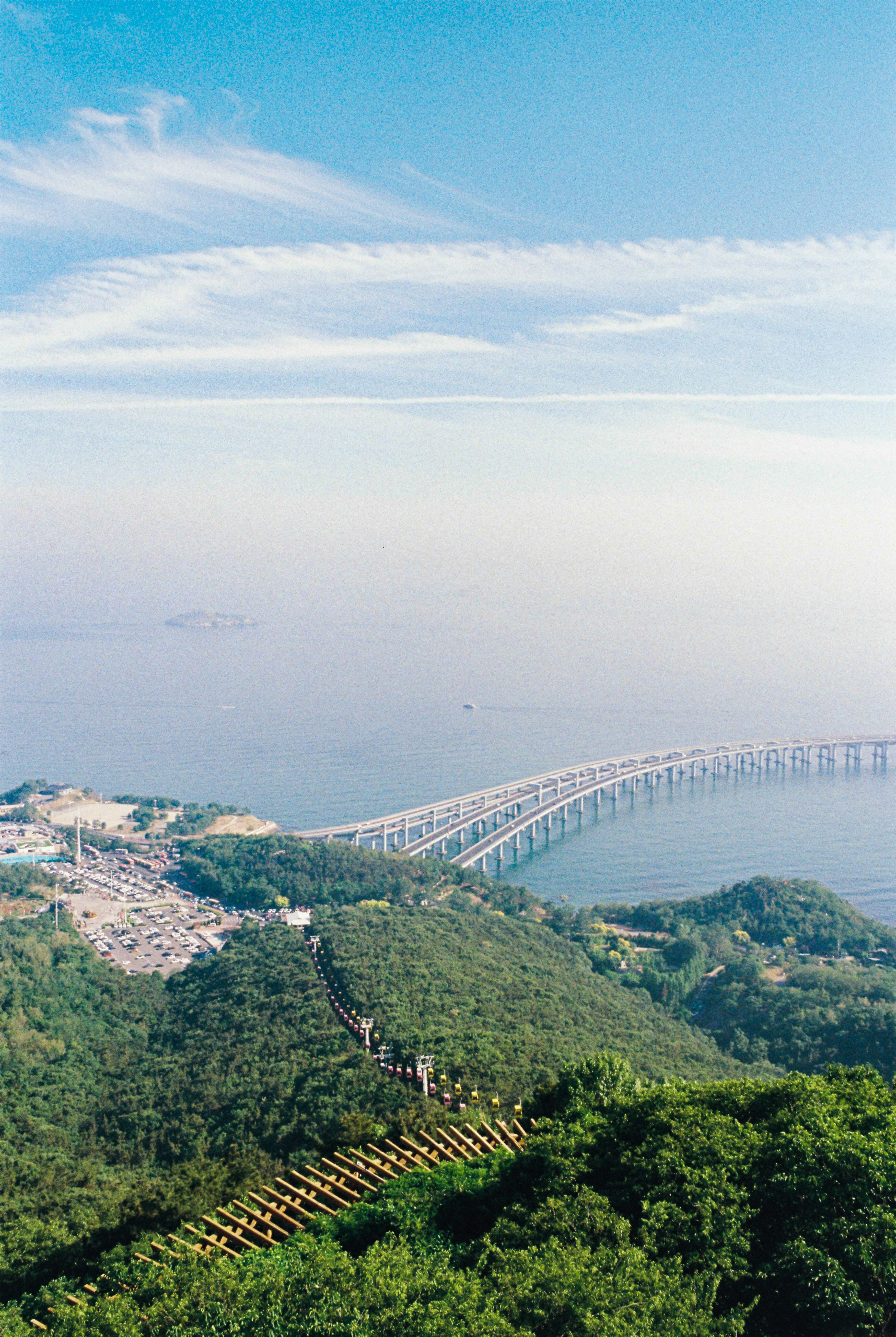 A winding bridge connects the lush green hills to the serene ocean, with distant islands visible on the horizon. The scene captures the harmony between nature and infrastructure.