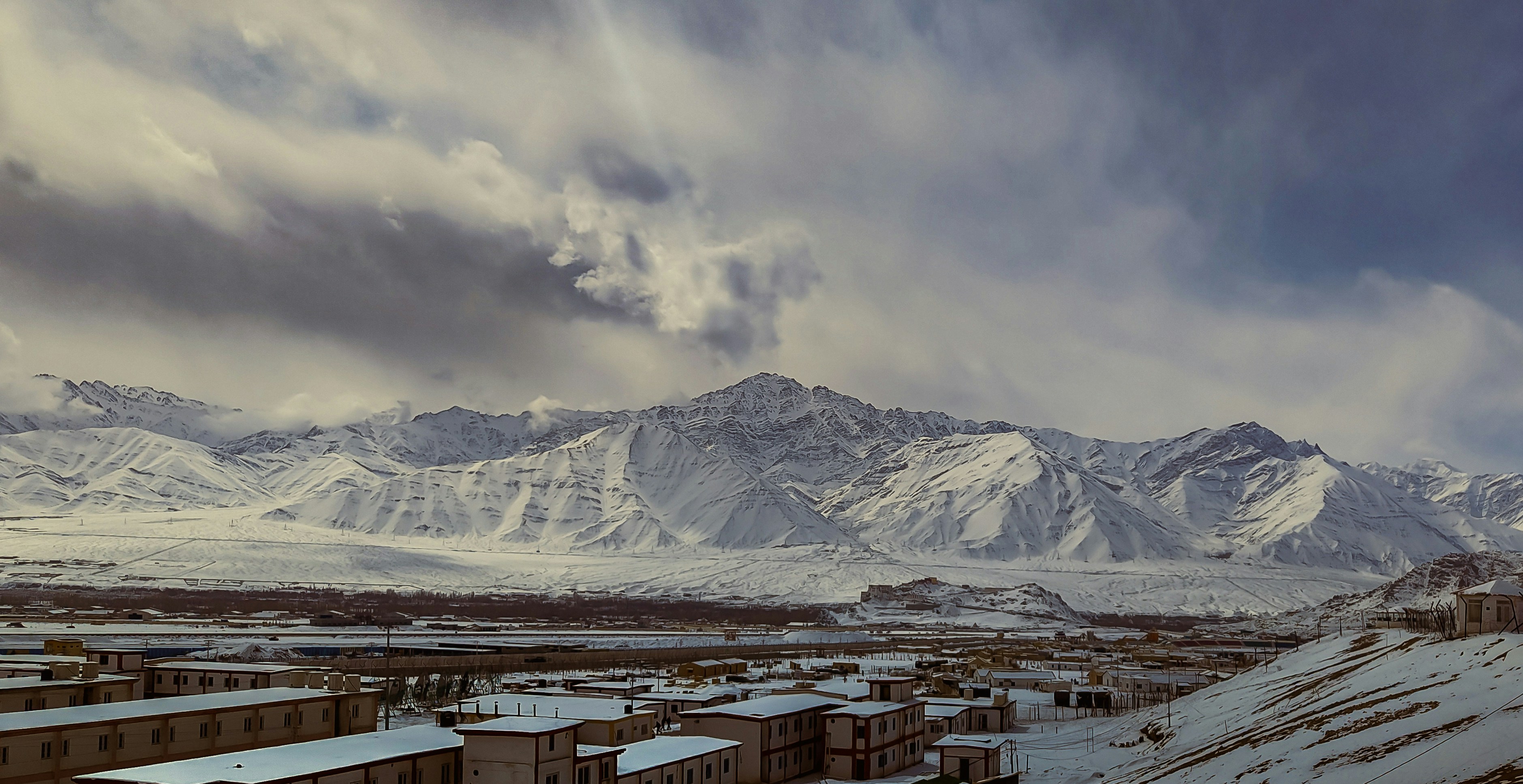 Snow-covered mountains loom over a village under cloudy skies.