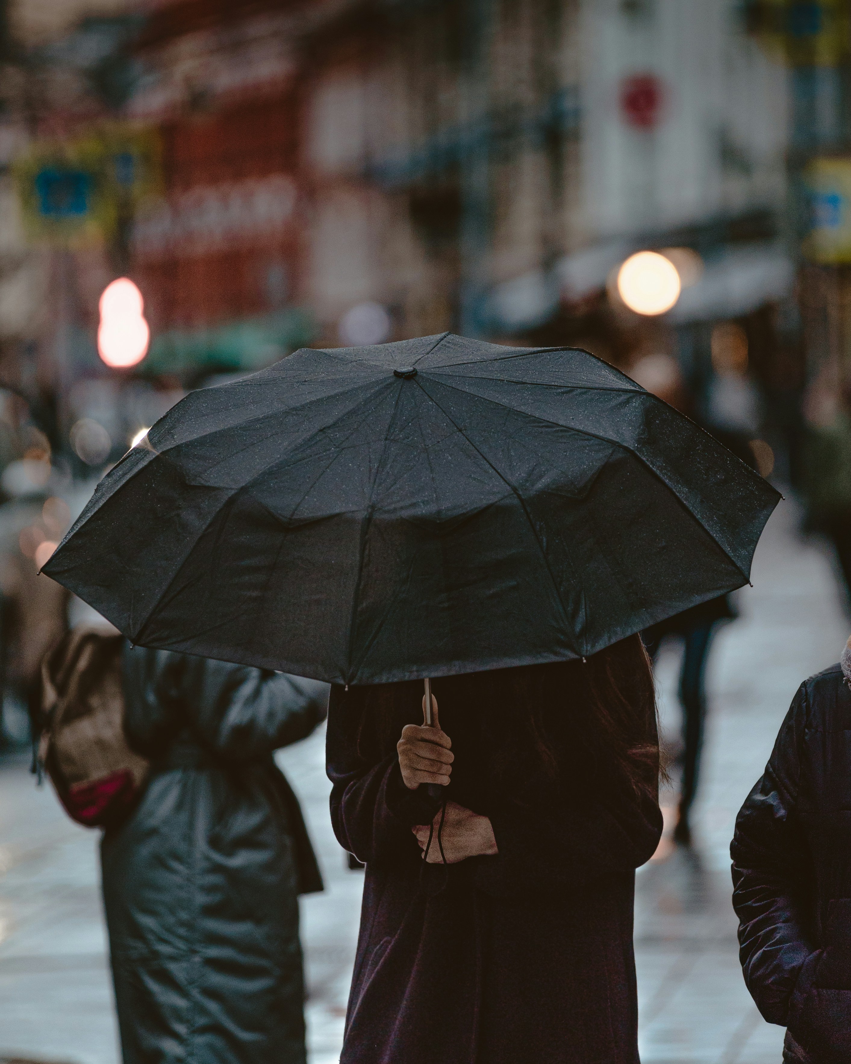 A figure holding a black umbrella stands amidst a blurred city street, evoking a sense of quiet introspection on a rainy day.