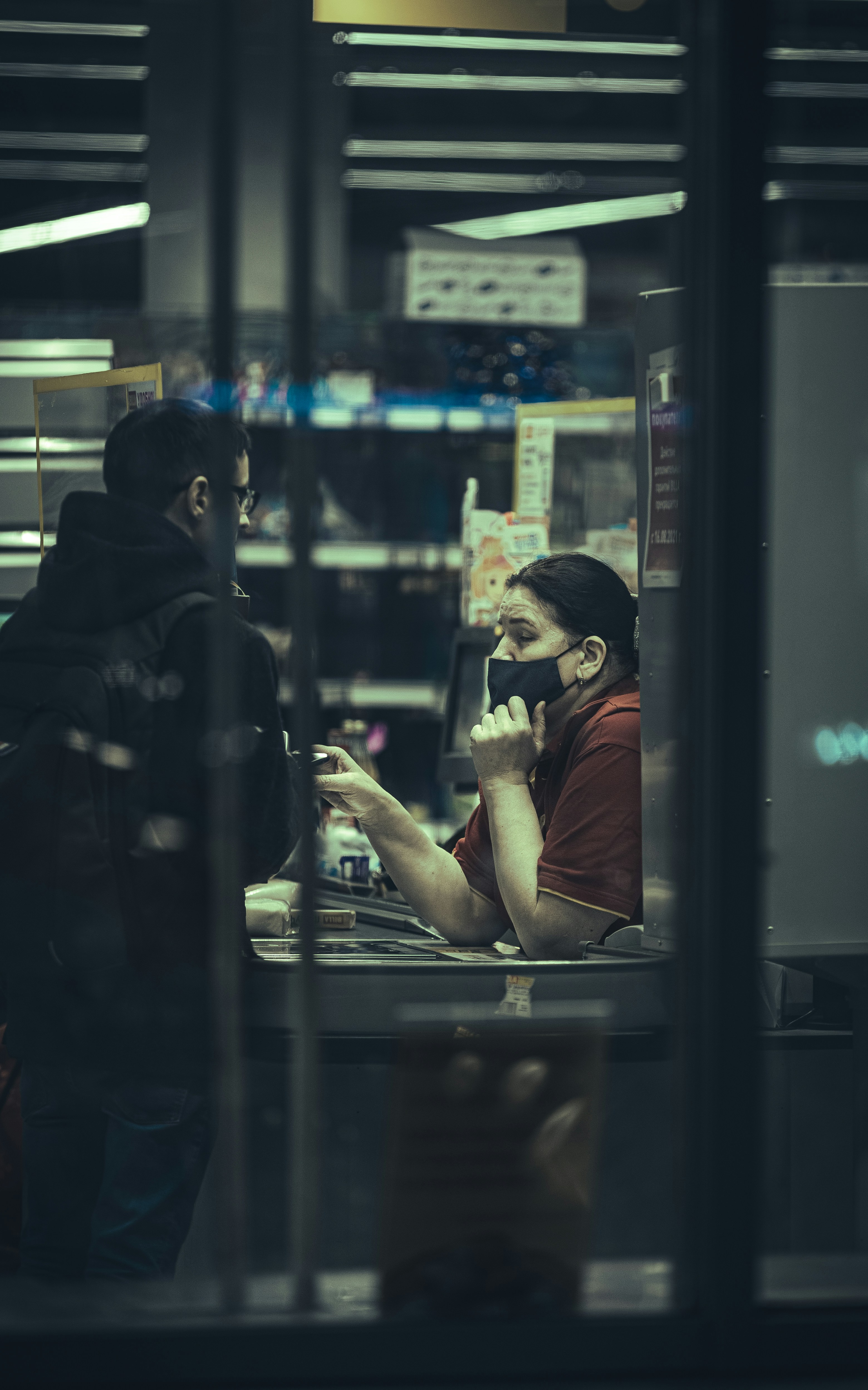 A customer engages in conversation with a cashier behind a glass partition in a dimly lit convenience store. The scene captures the essence of urban life during late hours.