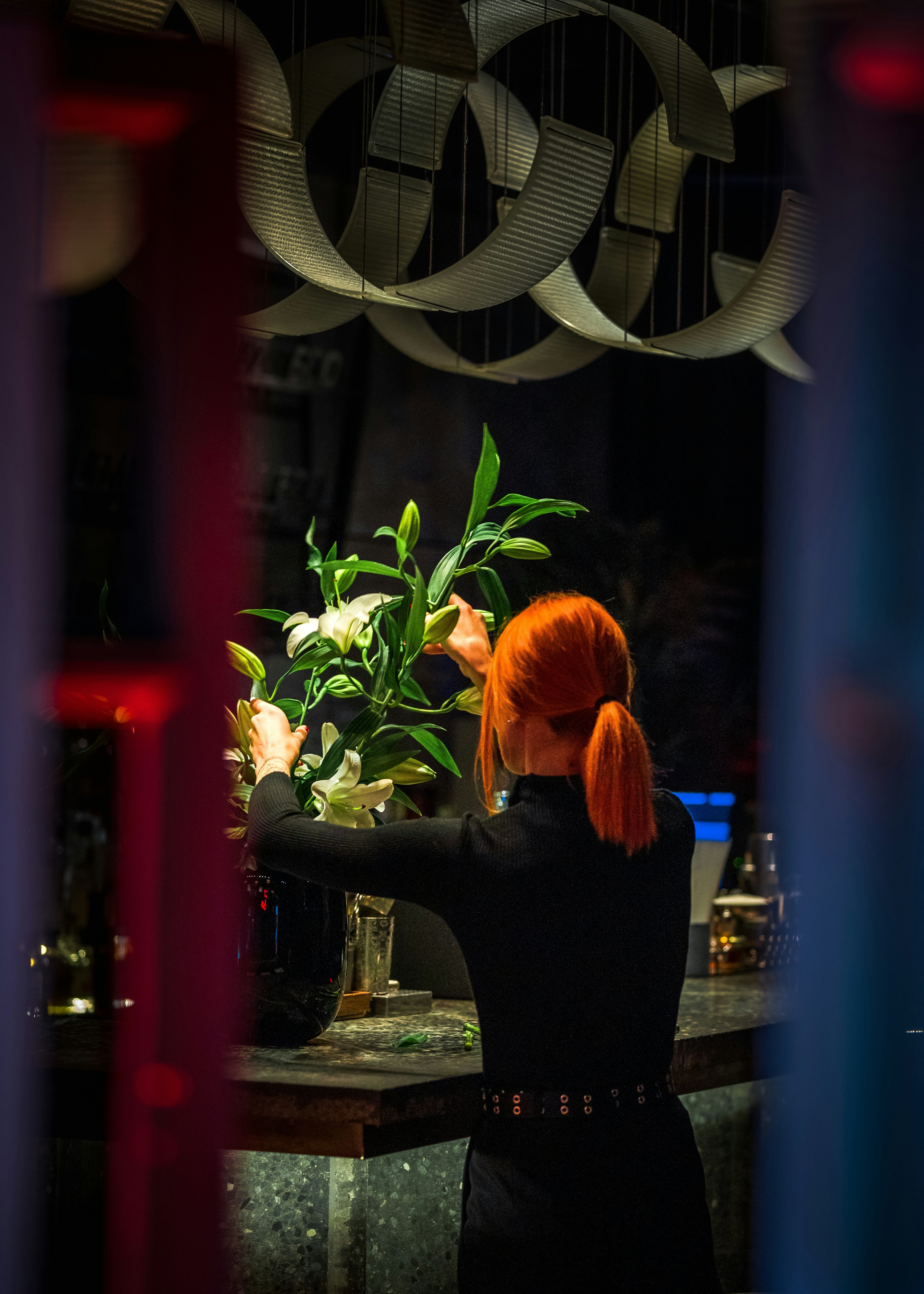 Red‑haired florist at a dimly lit bar arranging lilies under sculptural light