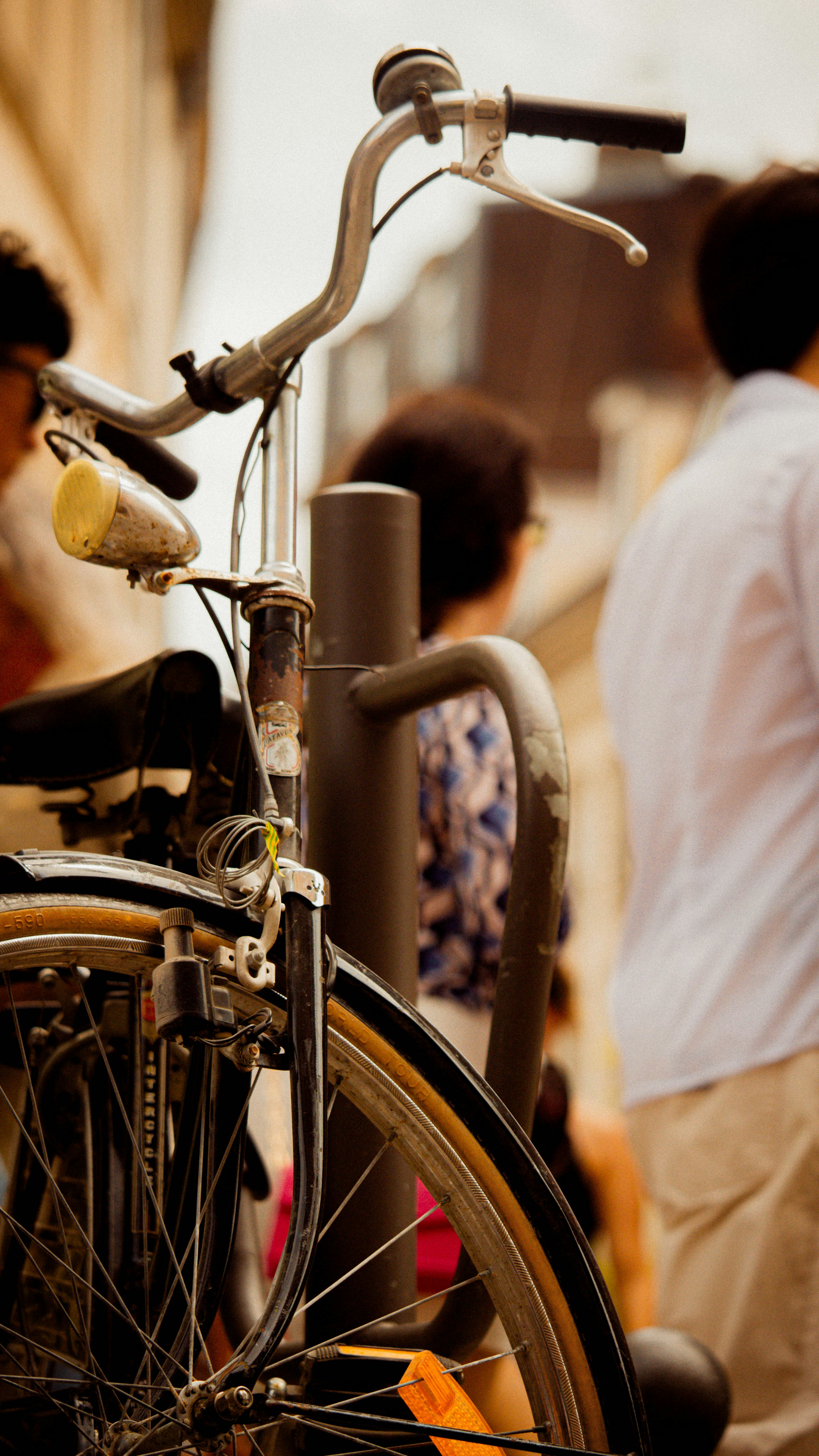 A vintage bicycle leans against a post, surrounded by blurred figures in a bustling street scene. The focus on the bike highlights its nostalgic charm amidst urban life.