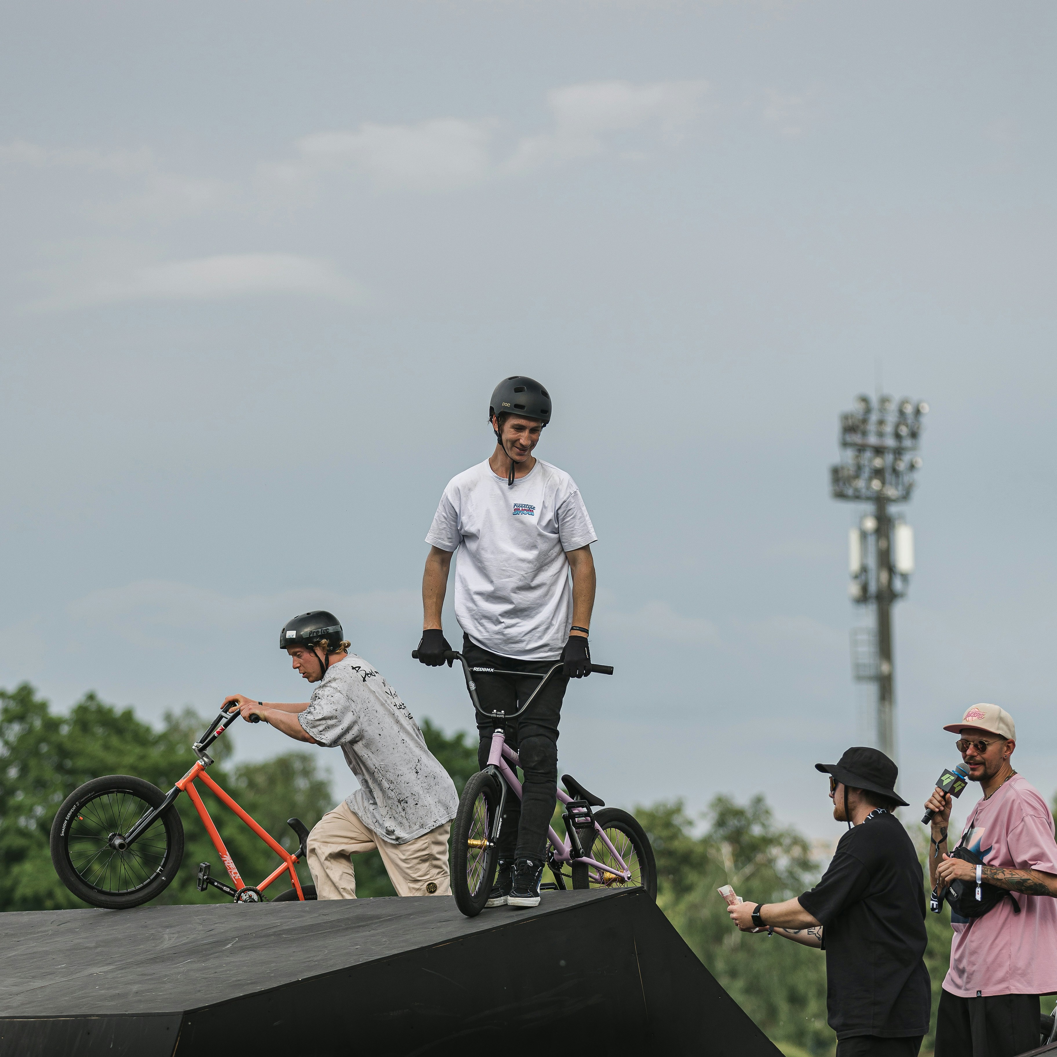 Three BMX riders performing tricks on a ramp at a skatepark, with spectators engaged in the background.