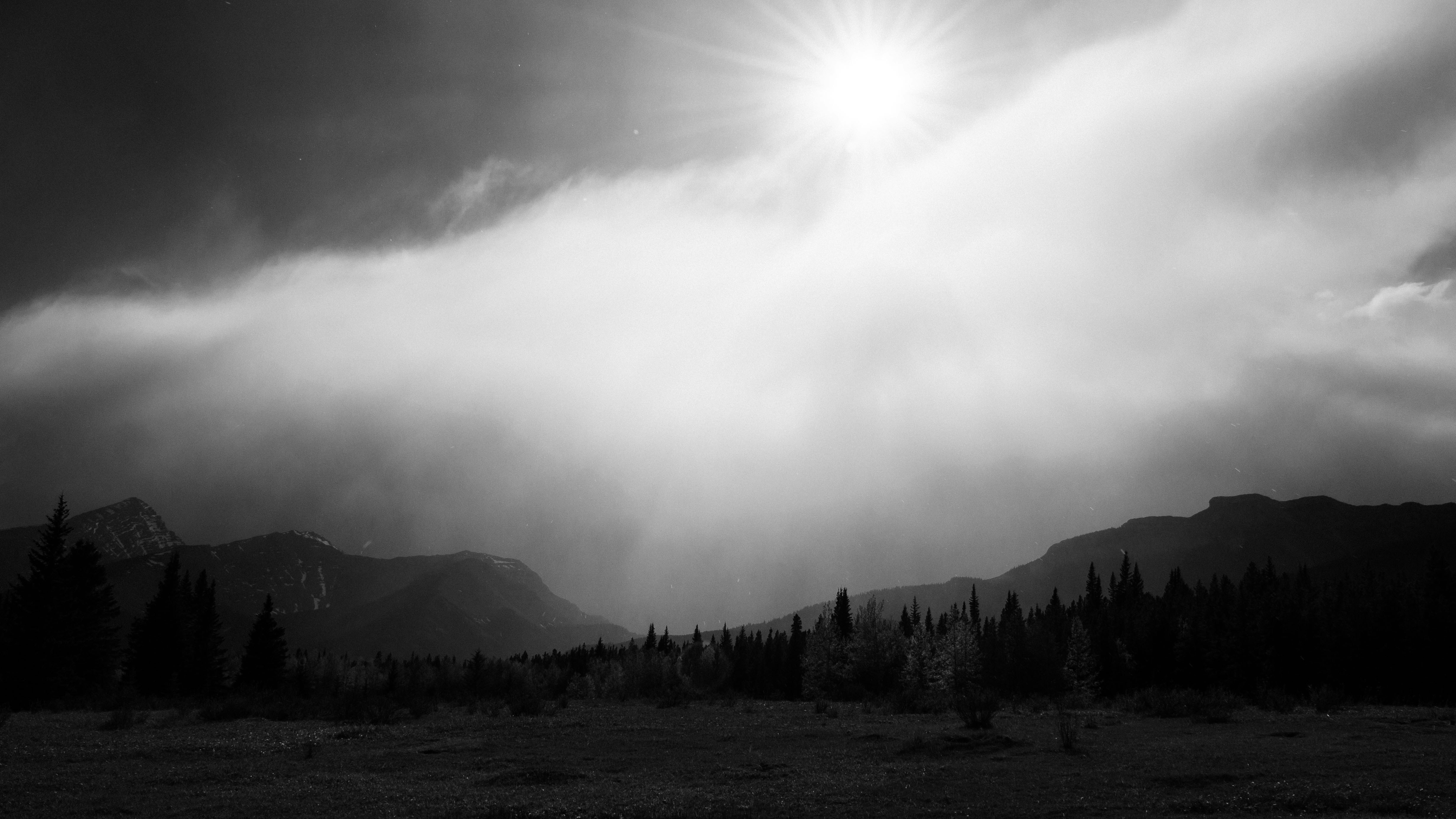 A striking black and white landscape capturing mountain silhouettes beneath a radiant sun and dynamic cloud formations, evoking a sense of grandeur and natural power. | Sunlight breaking through stormy clouds over mountains