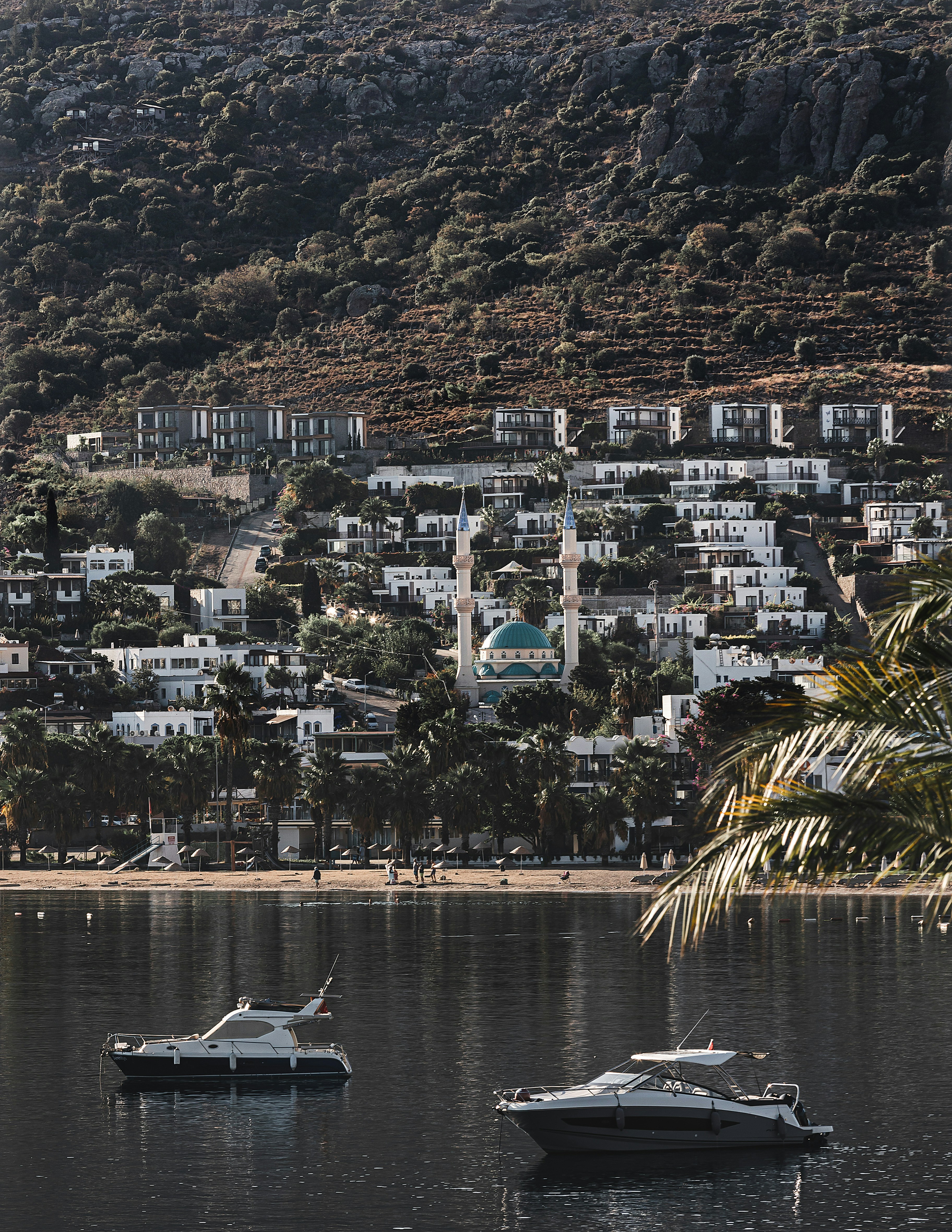 A tranquil coastal scene featuring boats anchored in still waters, framed by a hillside village and a prominent mosque. The setting reflects a serene blend of nature and architecture.