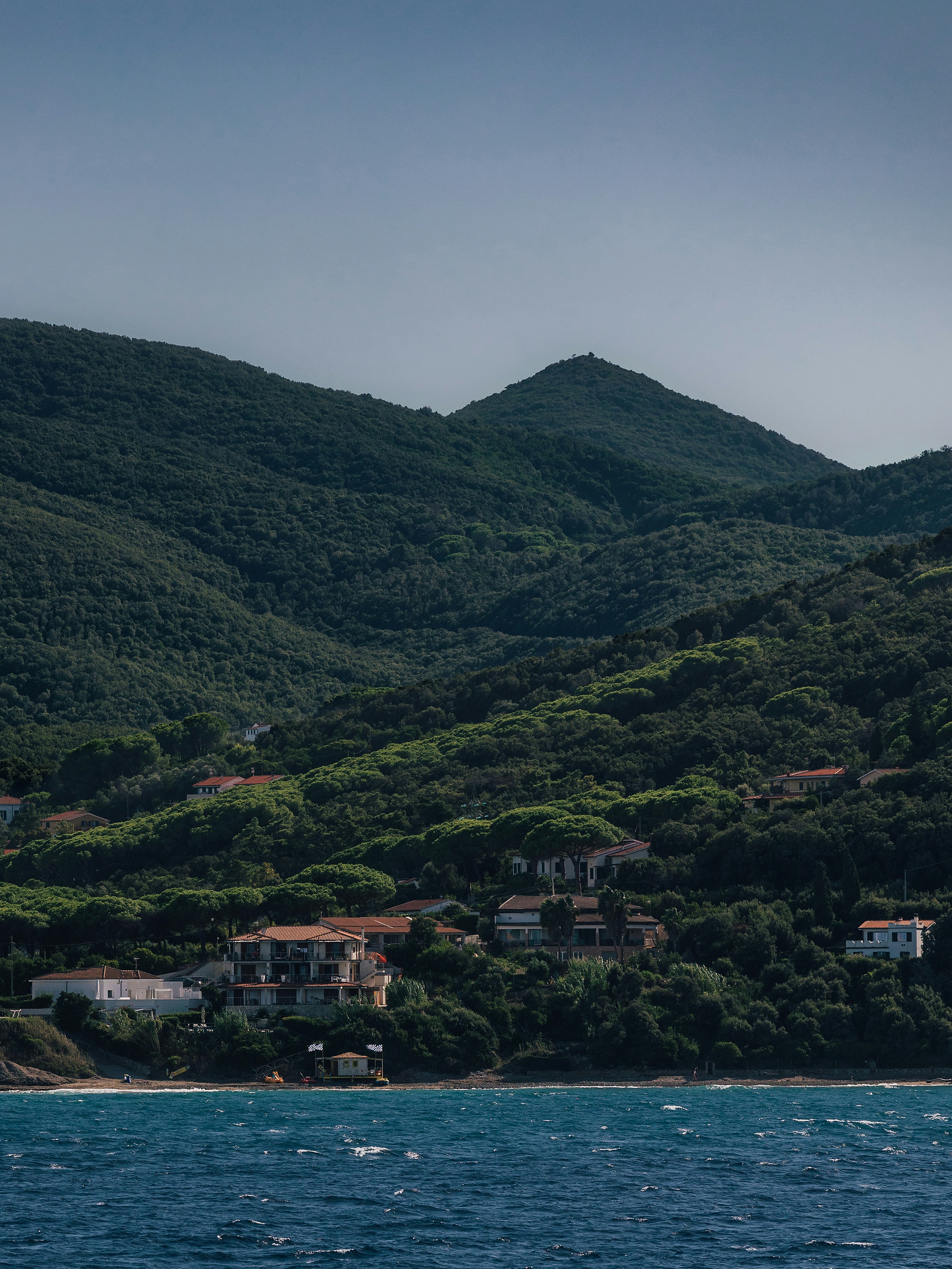 Lush green hills rise steeply behind a quaint coastal village, framed by the shimmering sea below.