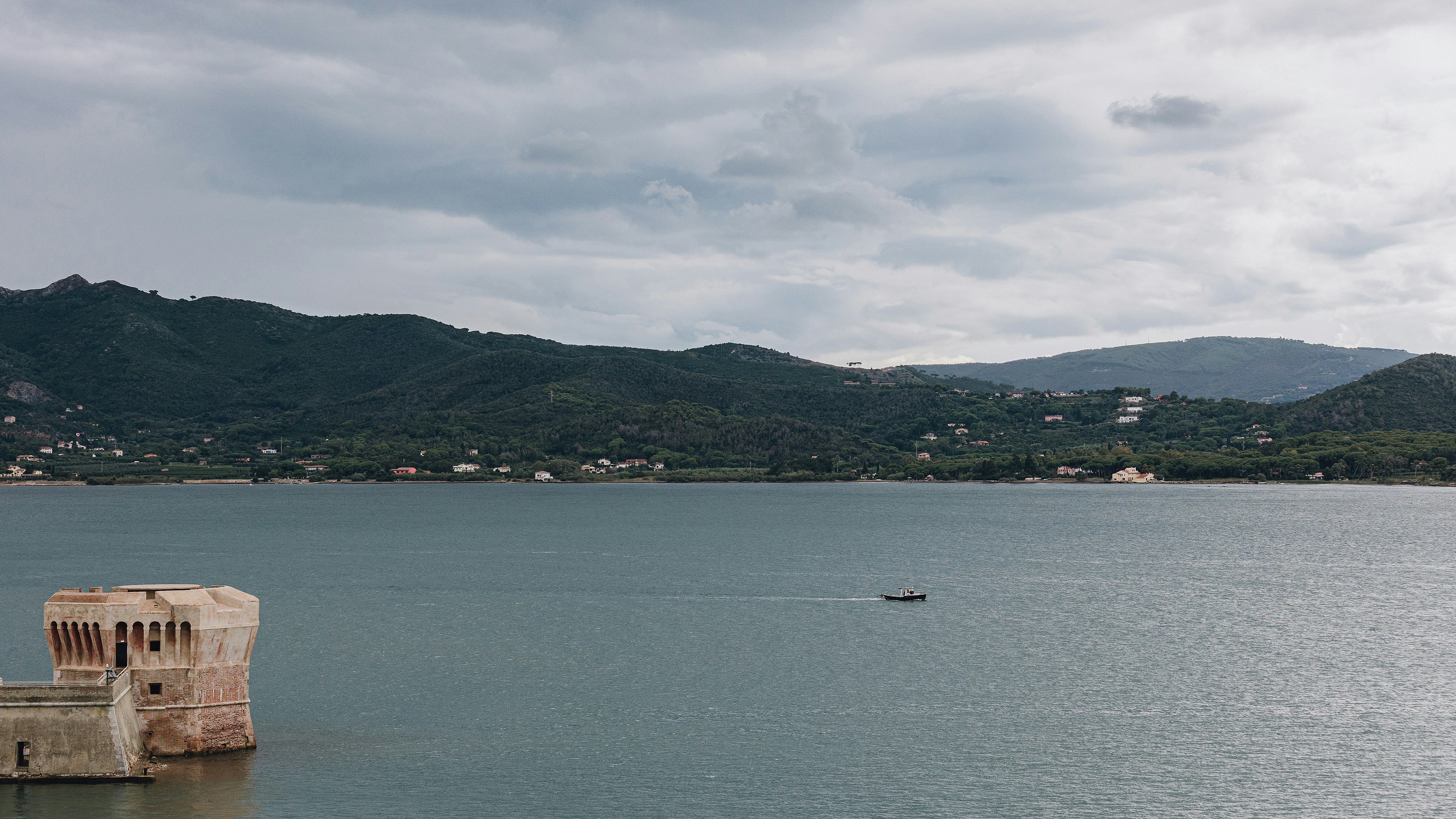 A tranquil view of a coastal landscape featuring a historic structure by the water, with a lone boat gliding across the surface. The surrounding hills add depth to the serene atmosphere.