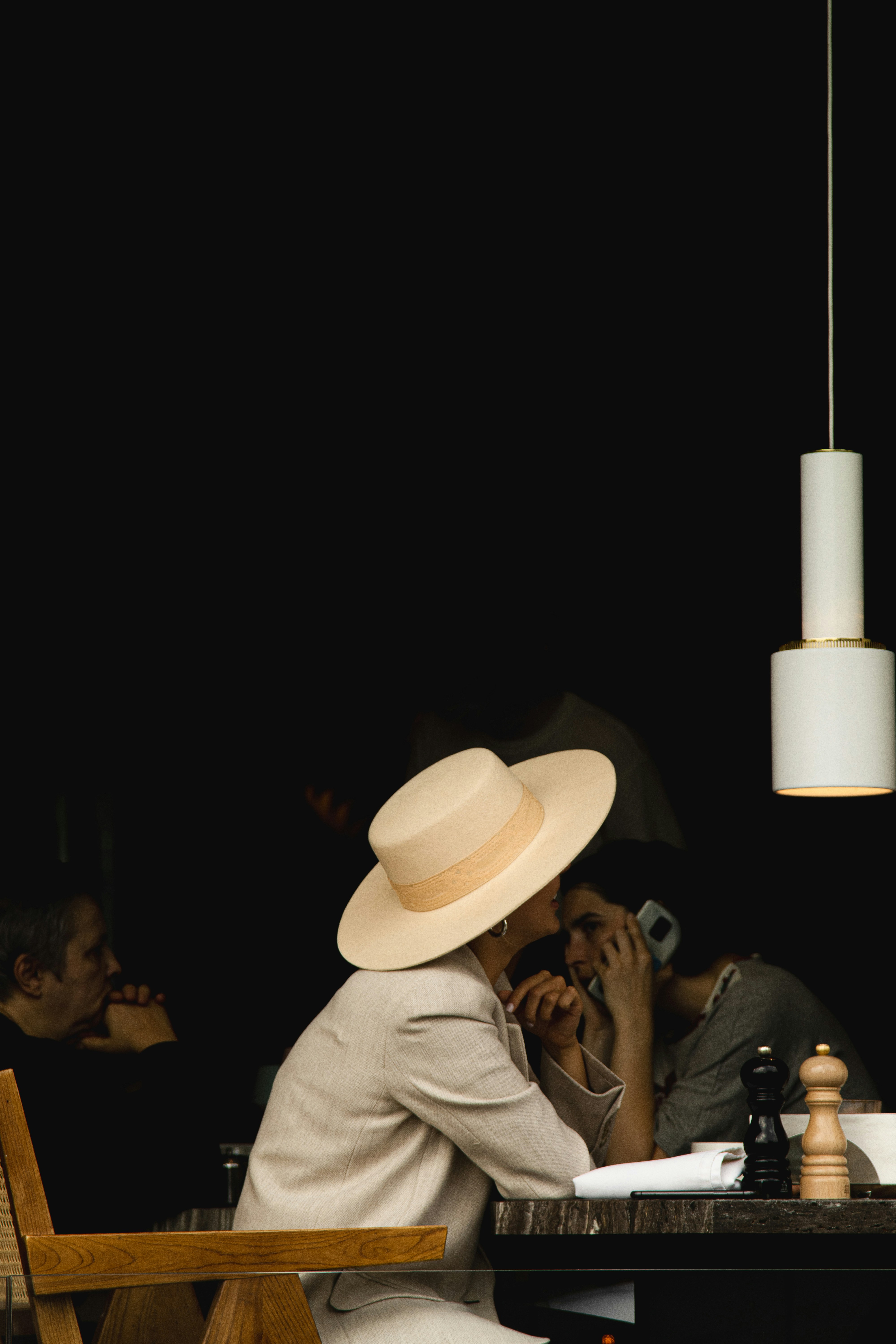 A woman in a wide-brimmed hat sits contemplatively at a café table, engaged in a phone conversation amidst a shadowy backdrop.