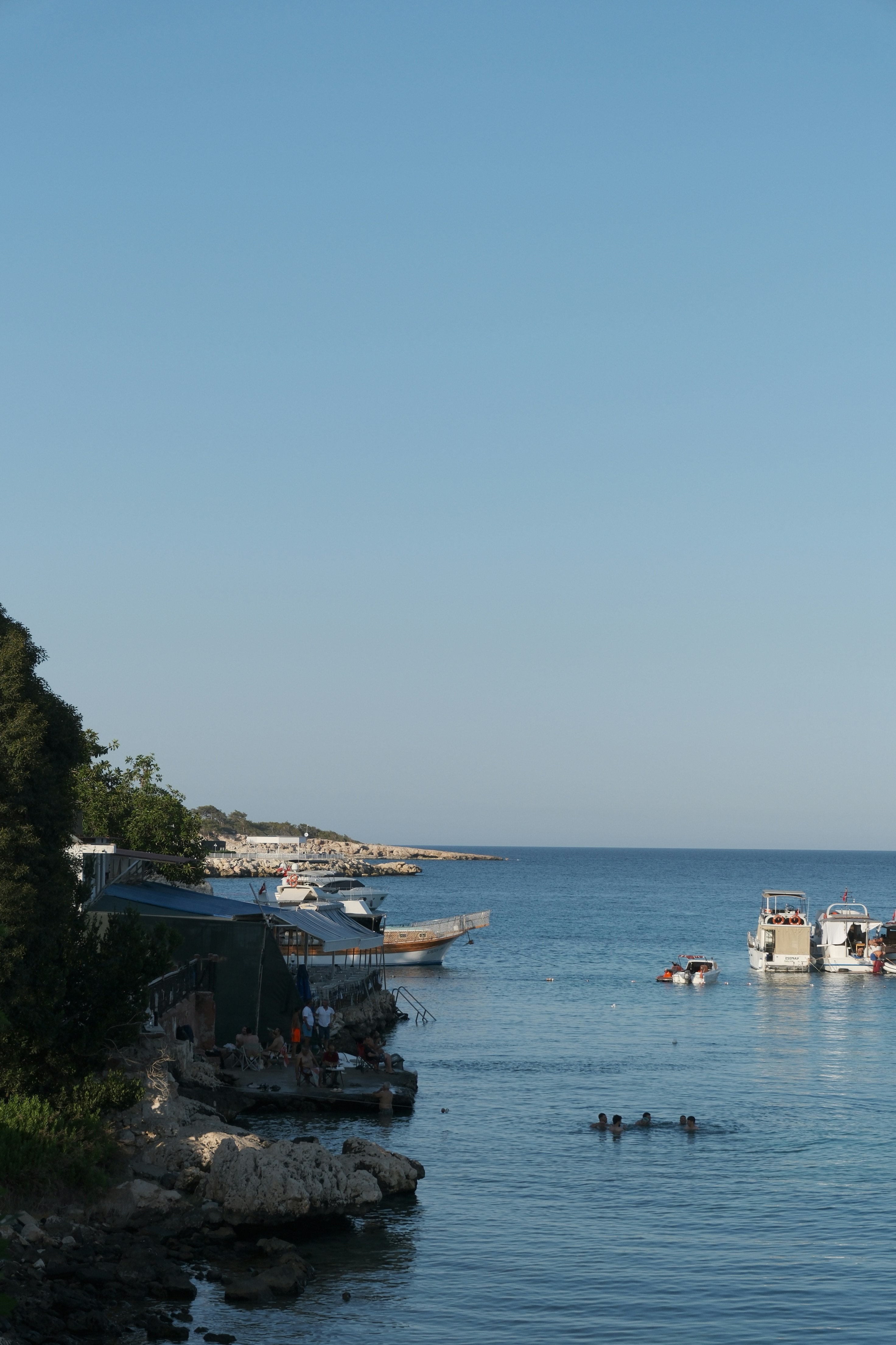Boats anchored in calm waters with swimmers enjoying the sun near a rocky shoreline. Lush greenery frames the scene.