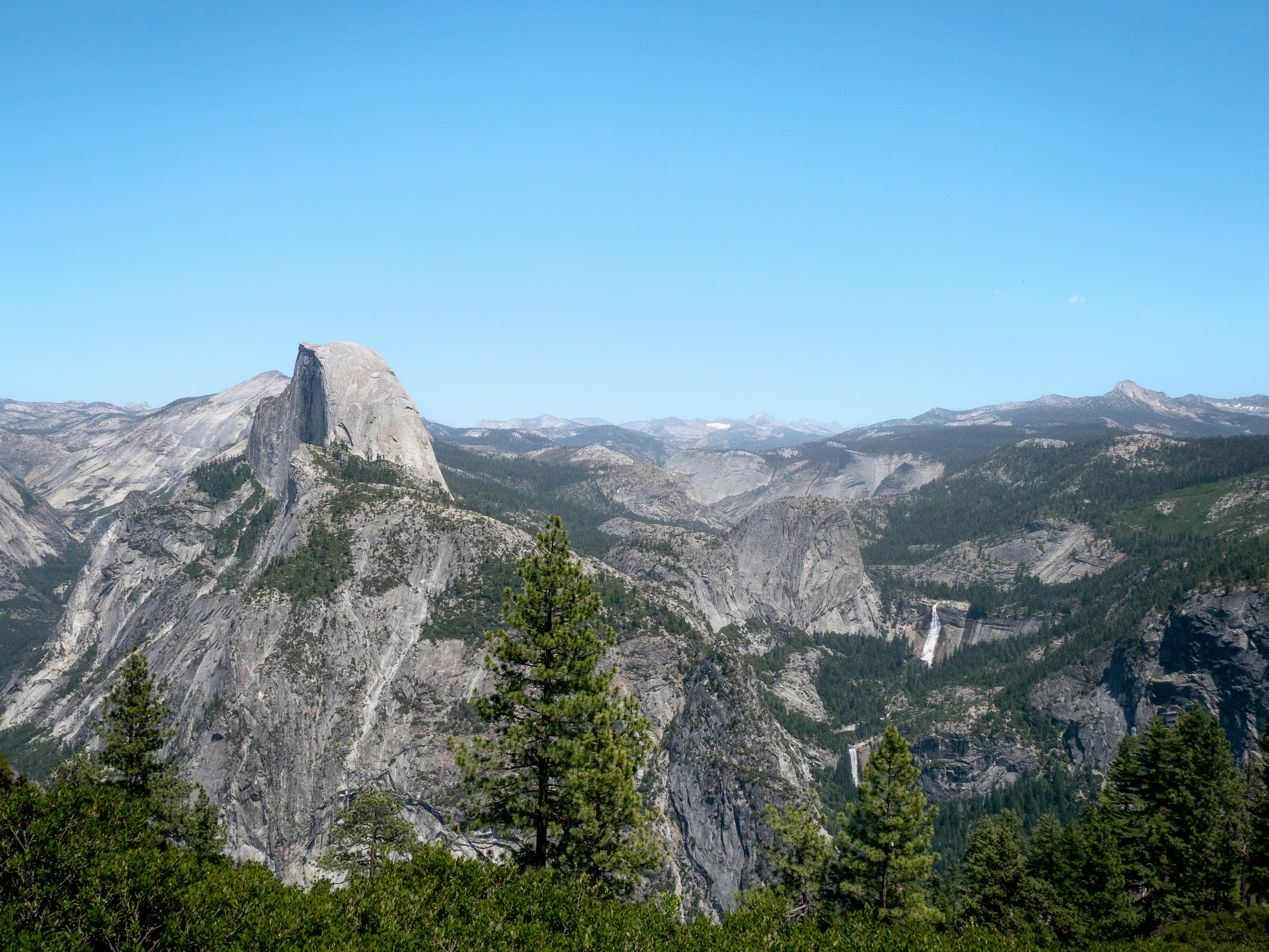 A panoramic view of Half Dome rising majestically above the Yosemite Valley, framed by lush green trees and distant mountain ranges.