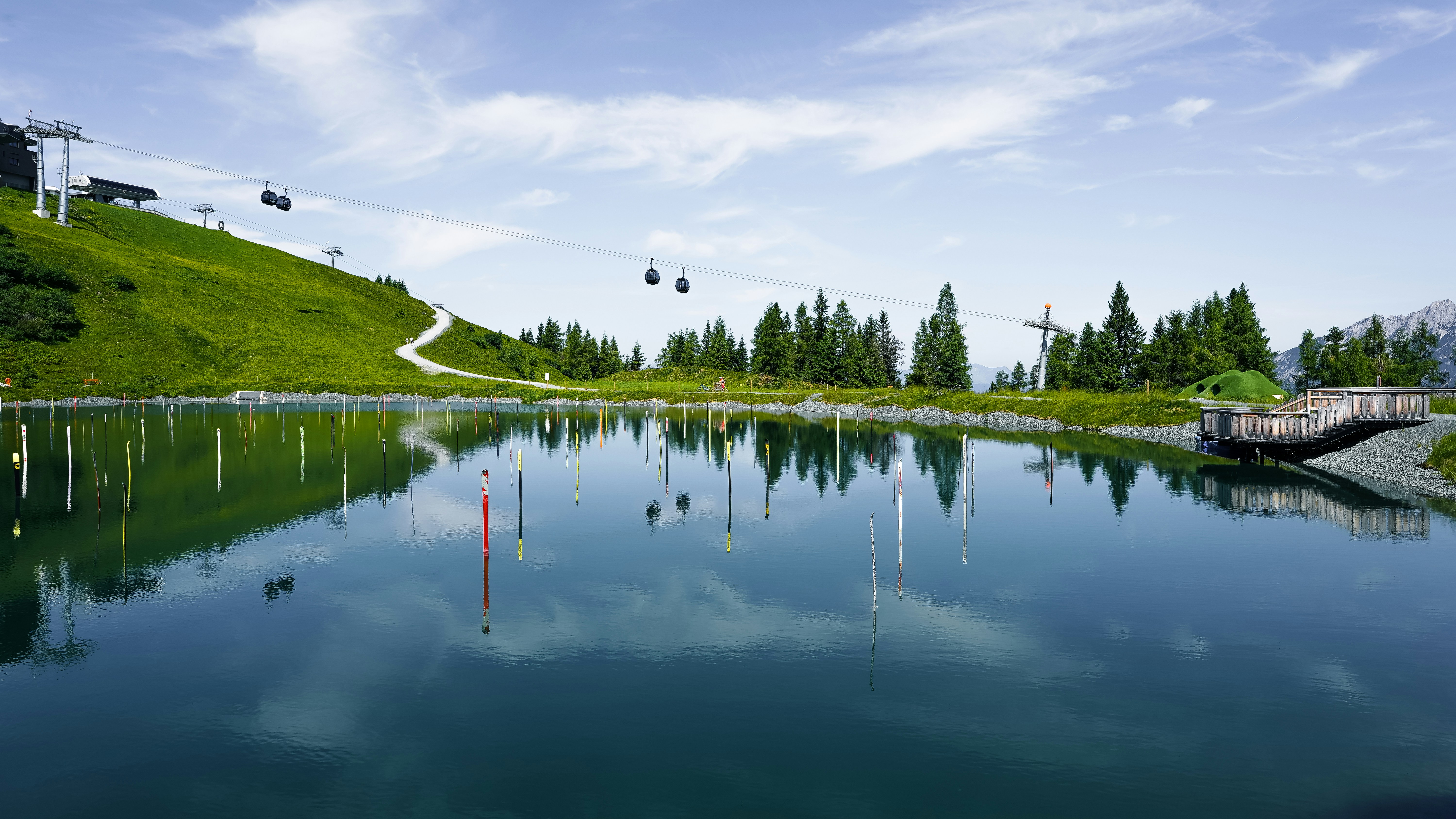 Serene alpine lake reflecting the surrounding greenery and cable cars overhead, with colorful poles marking the water's edge.