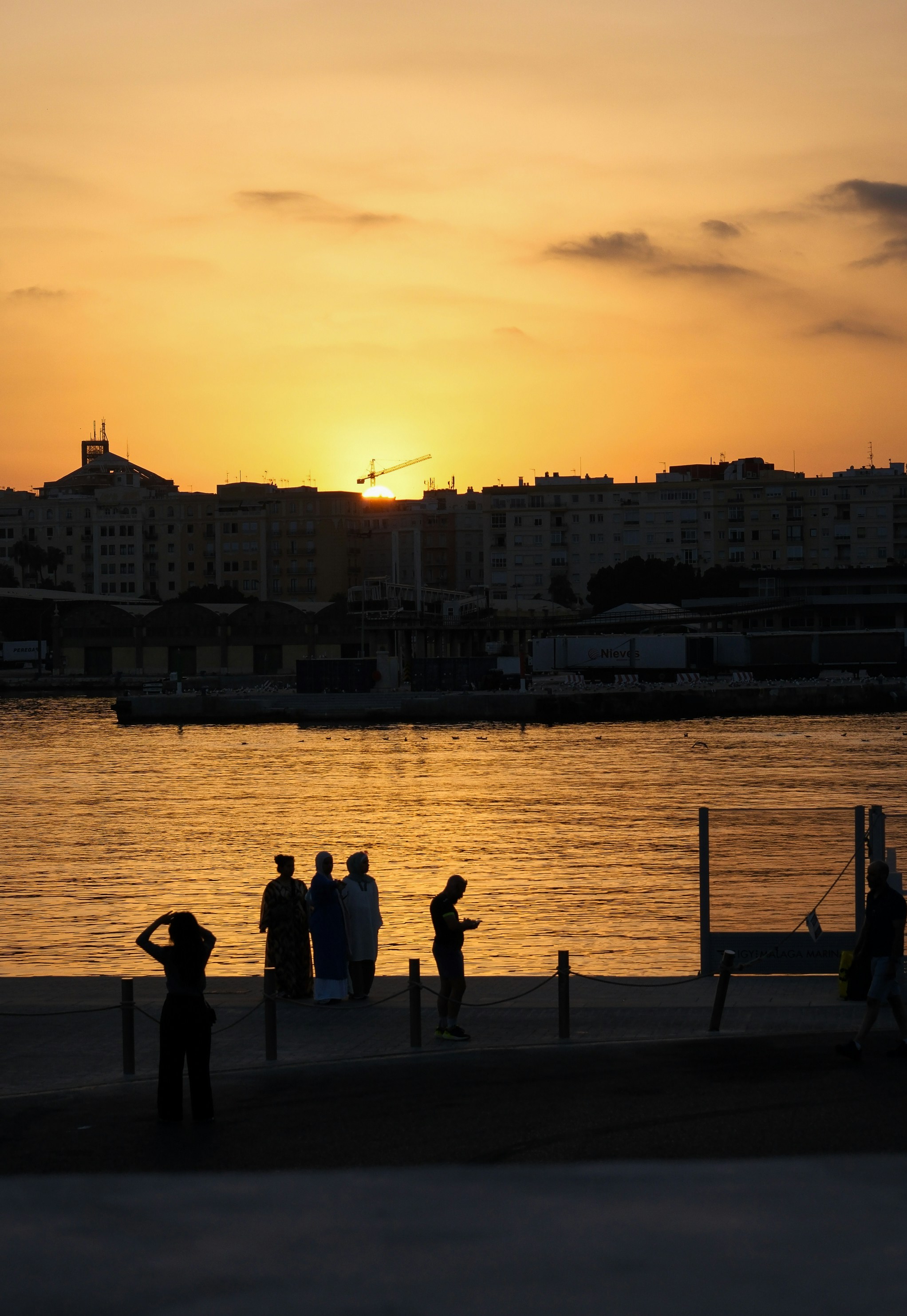 Silhouetted figures gather along a waterfront as the sun sets, casting a warm glow over the cityscape. The scene captures a tranquil evening atmosphere.