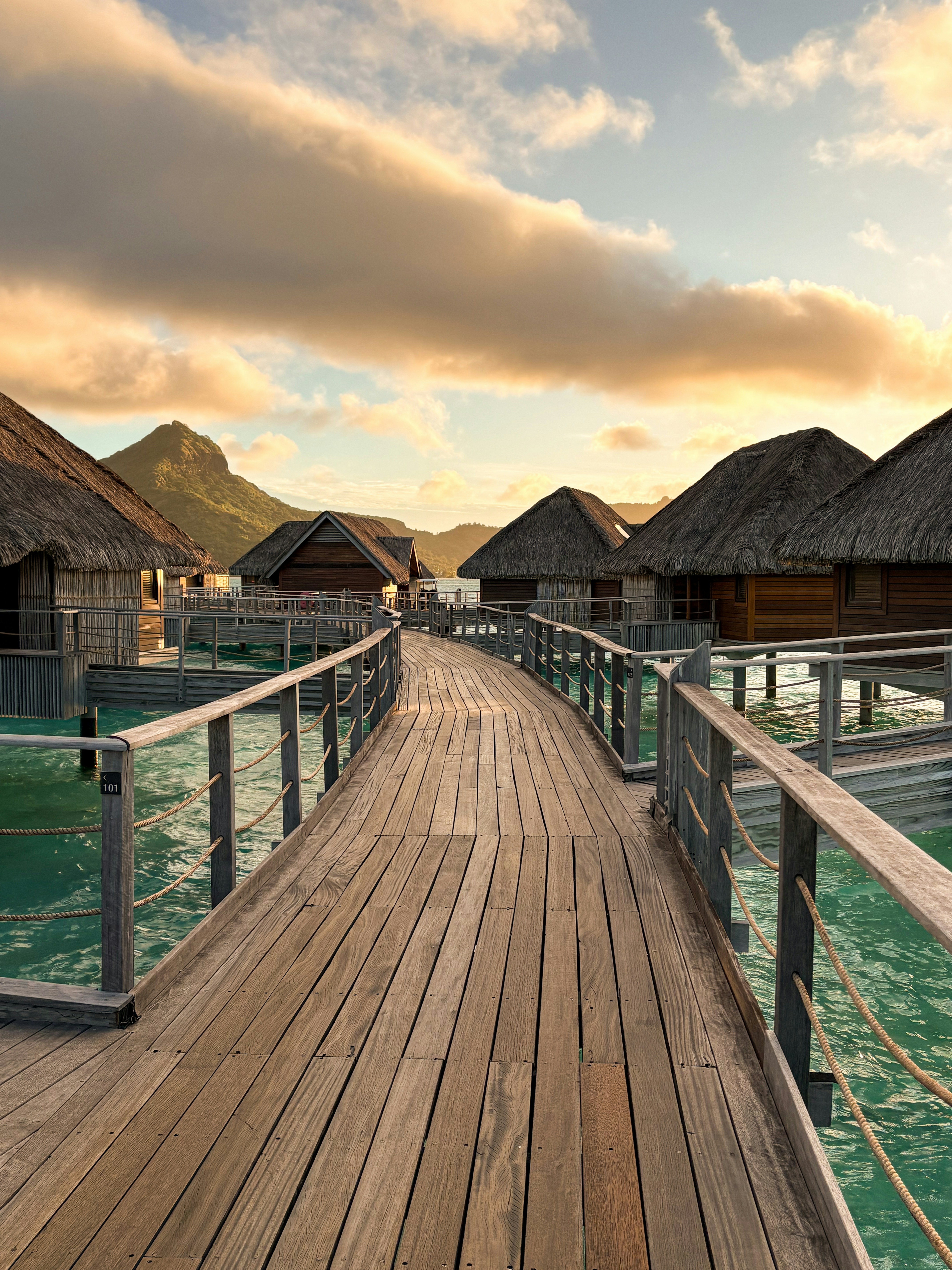Wooden walkway leading through overwater bungalows, framed by lush mountains and a colorful sky at sunset.