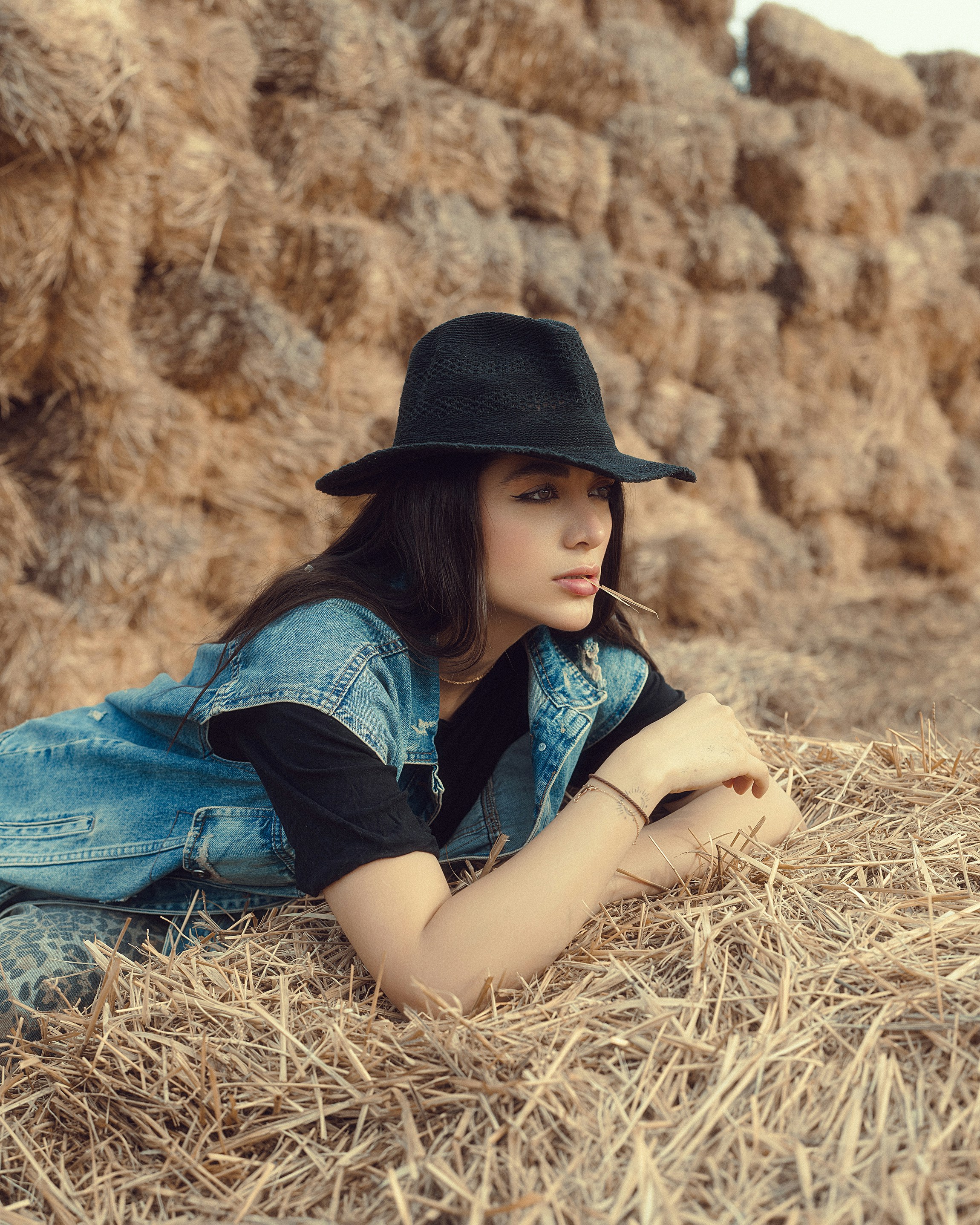Young woman reclining on straw bales, wearing a denim vest and black hat, with a thoughtful expression. The rustic backdrop enhances the serene atmosphere.
