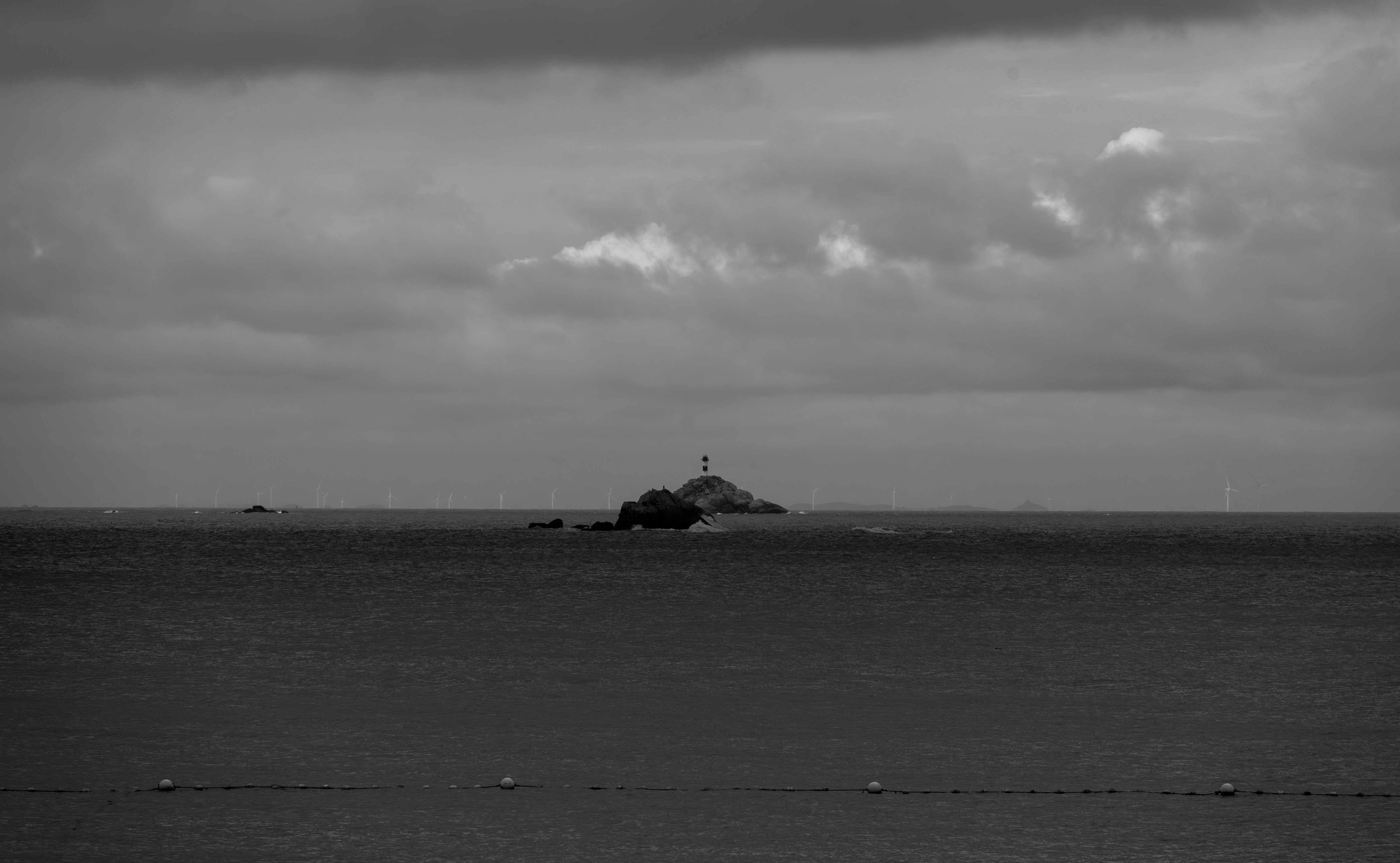 A solitary lighthouse stands on a rocky islet amidst a vast, tranquil sea under a cloudy sky. The scene is rendered in black and white, enhancing the mood of isolation.