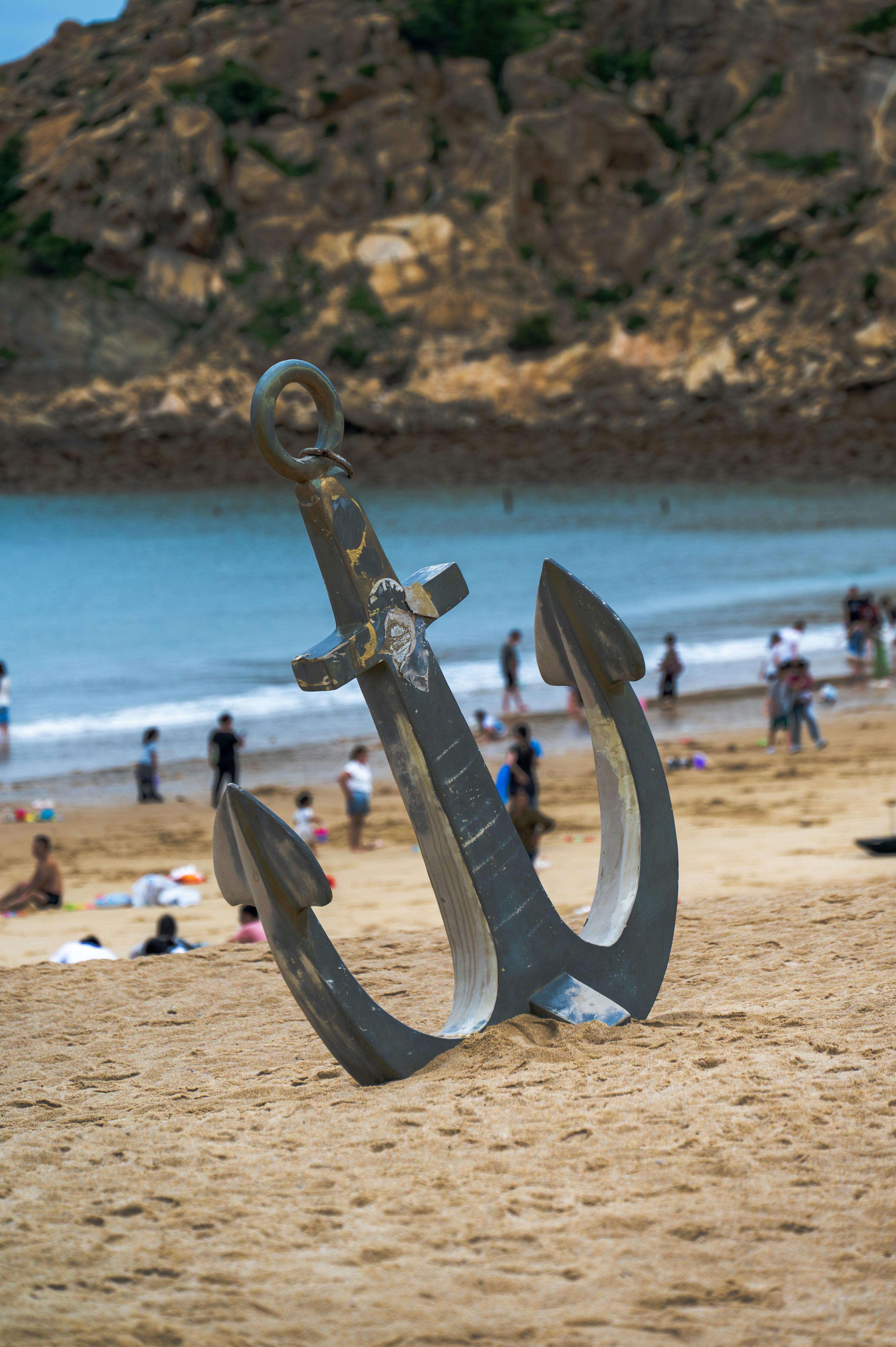 A large metallic anchor stands prominently on a sandy beach, with beachgoers enjoying the sun and surf in the background.