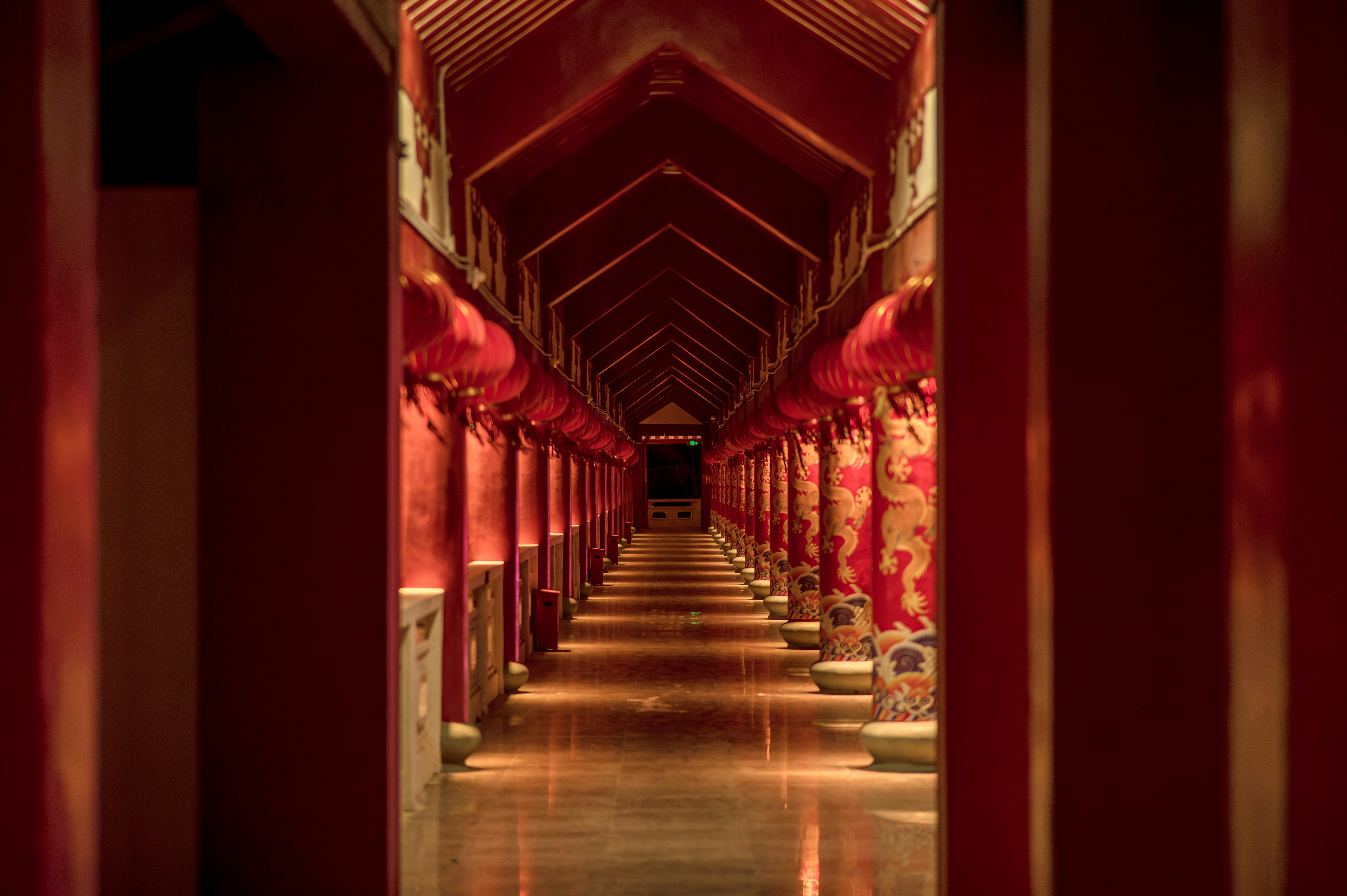 A luminous corridor adorned with rich red hues and intricate designs, leading to a distant focal point. The atmosphere evokes a sense of history and reverence.