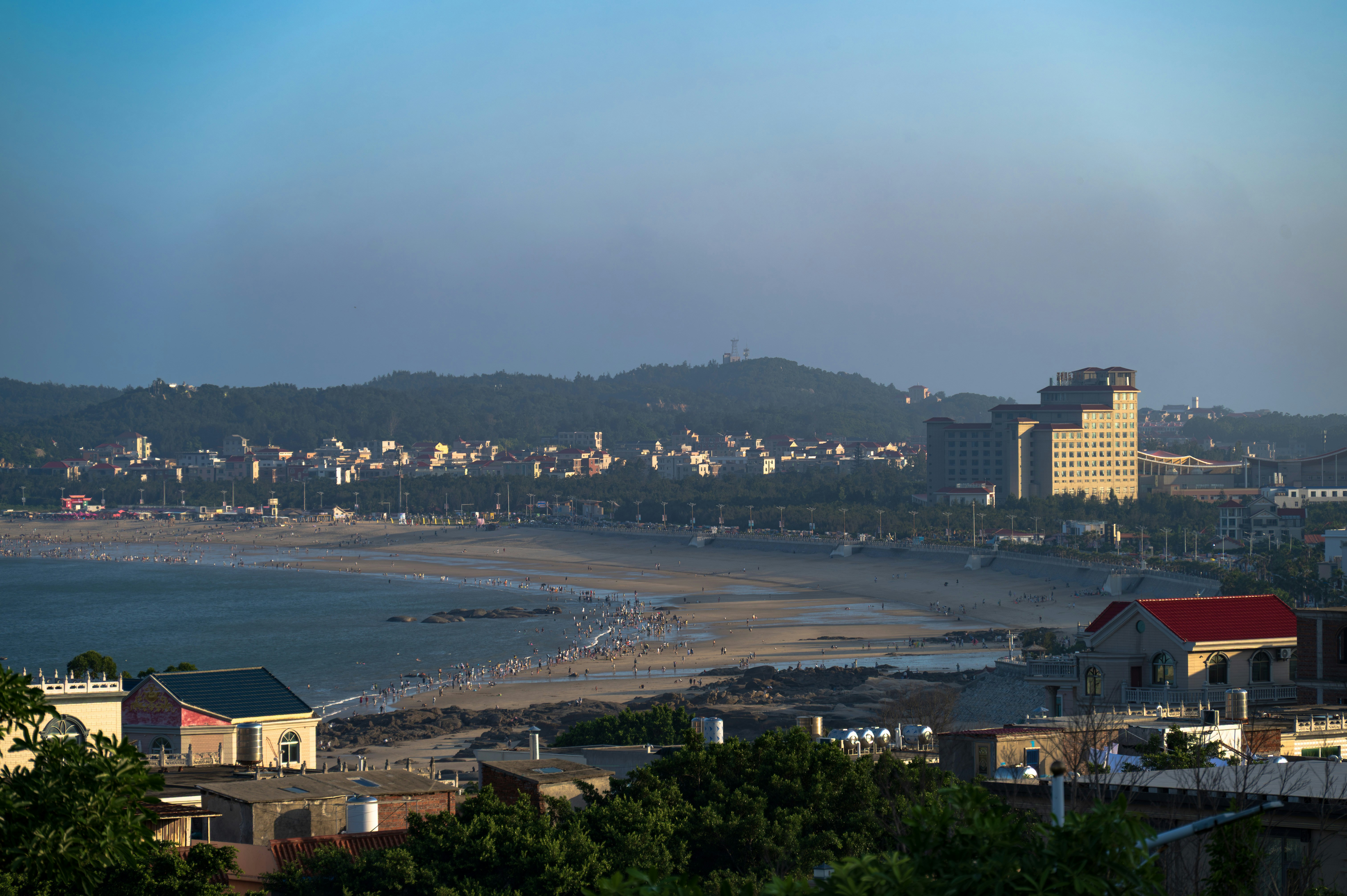 Expansive view of a beachside town with people strolling along the shore, framed by distant hills and a soft evening sky.