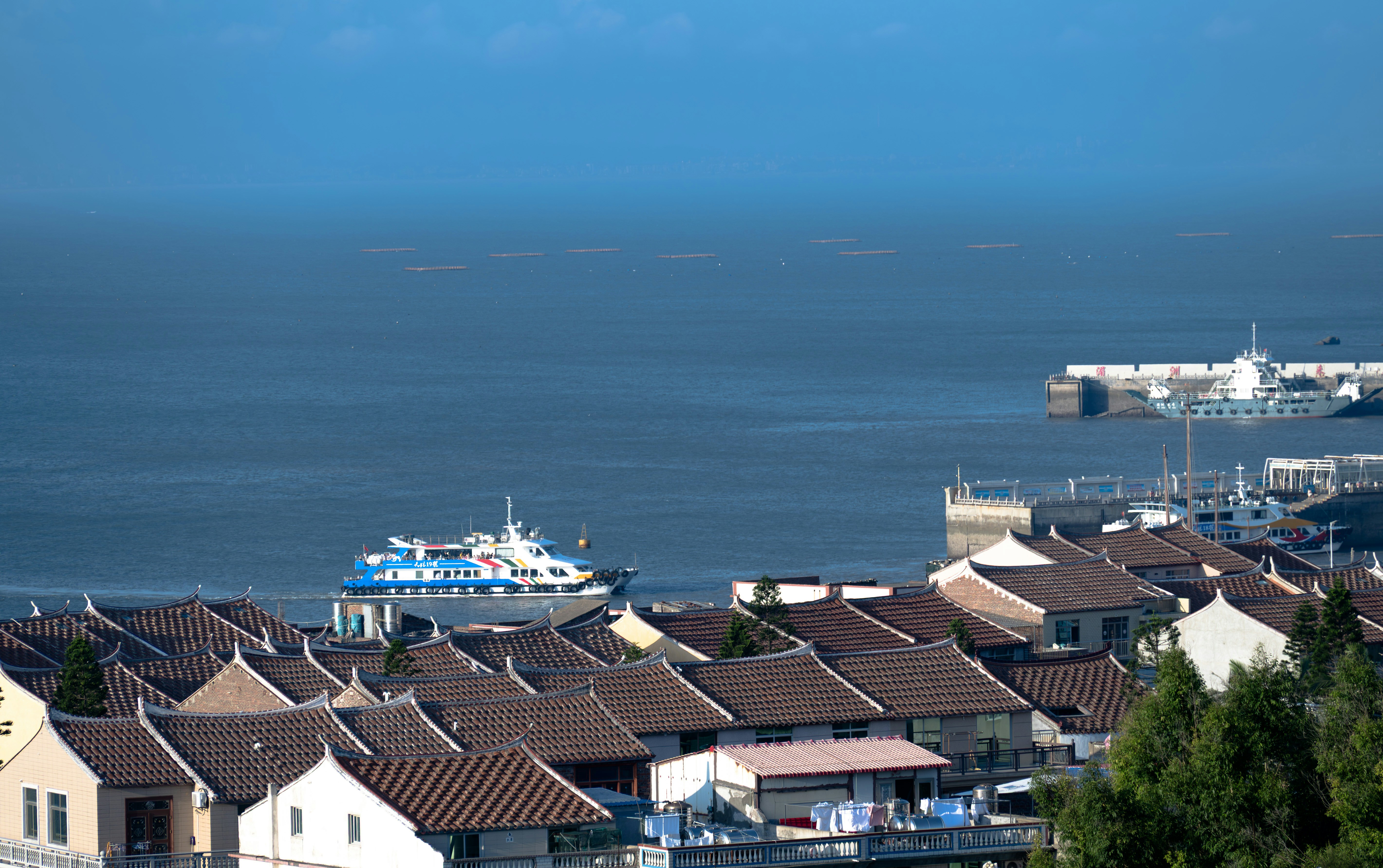 Aerial view of a coastal town with traditional rooftops and a harbor featuring boats docked at the pier. The calm sea stretches into the distance.