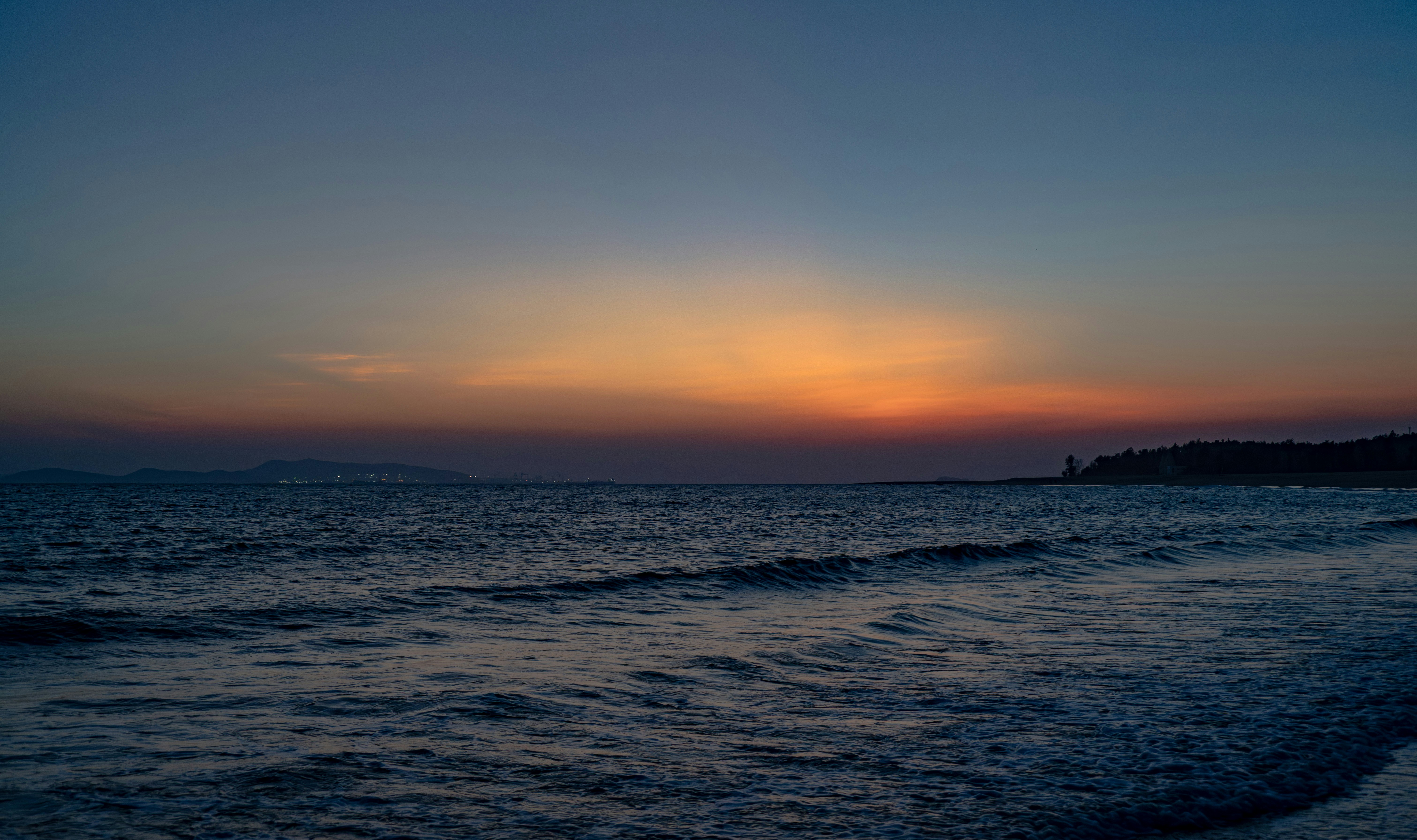 Gentle waves lap against the shore under a gradient sky transitioning from dusk to night. The distant lights of a coastal city twinkle on the horizon.