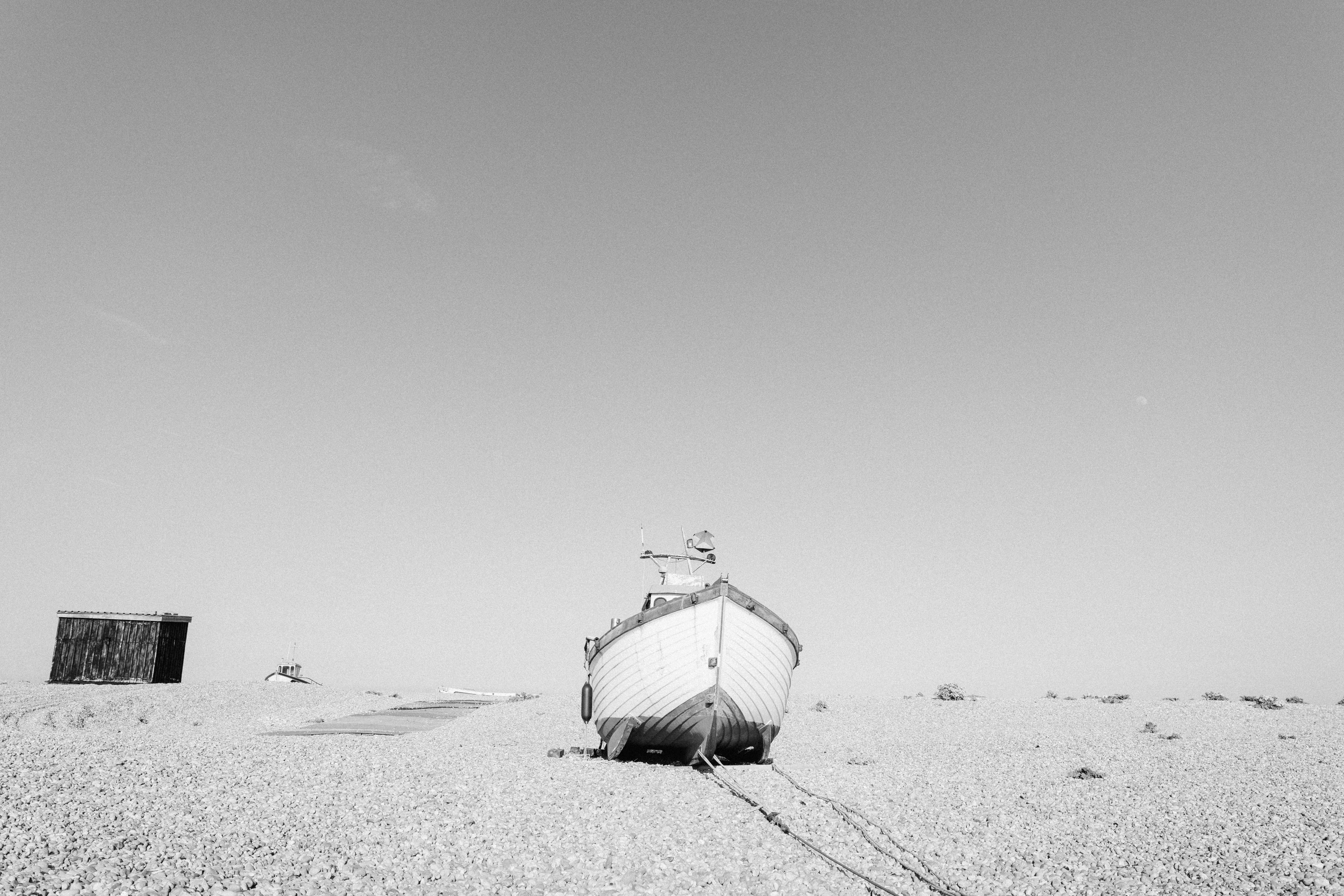 A weathered boat rests on a pebble-strewn beach under a clear sky, evoking a sense of solitude. A distant shed adds context to the tranquil scene.