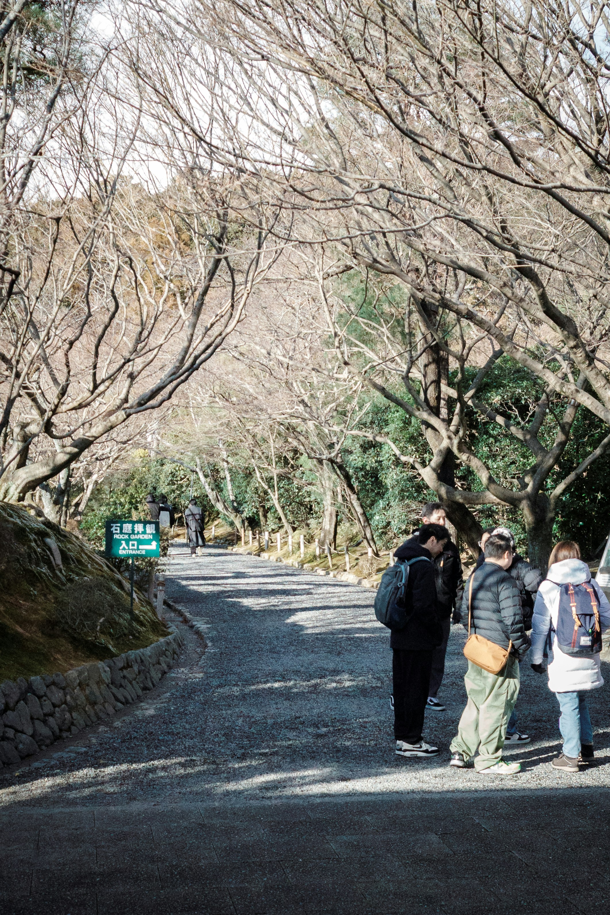 Visitors gather along a gravel path lined with bare trees, leading through a tranquil landscape. A sign indicates the entrance to the area.