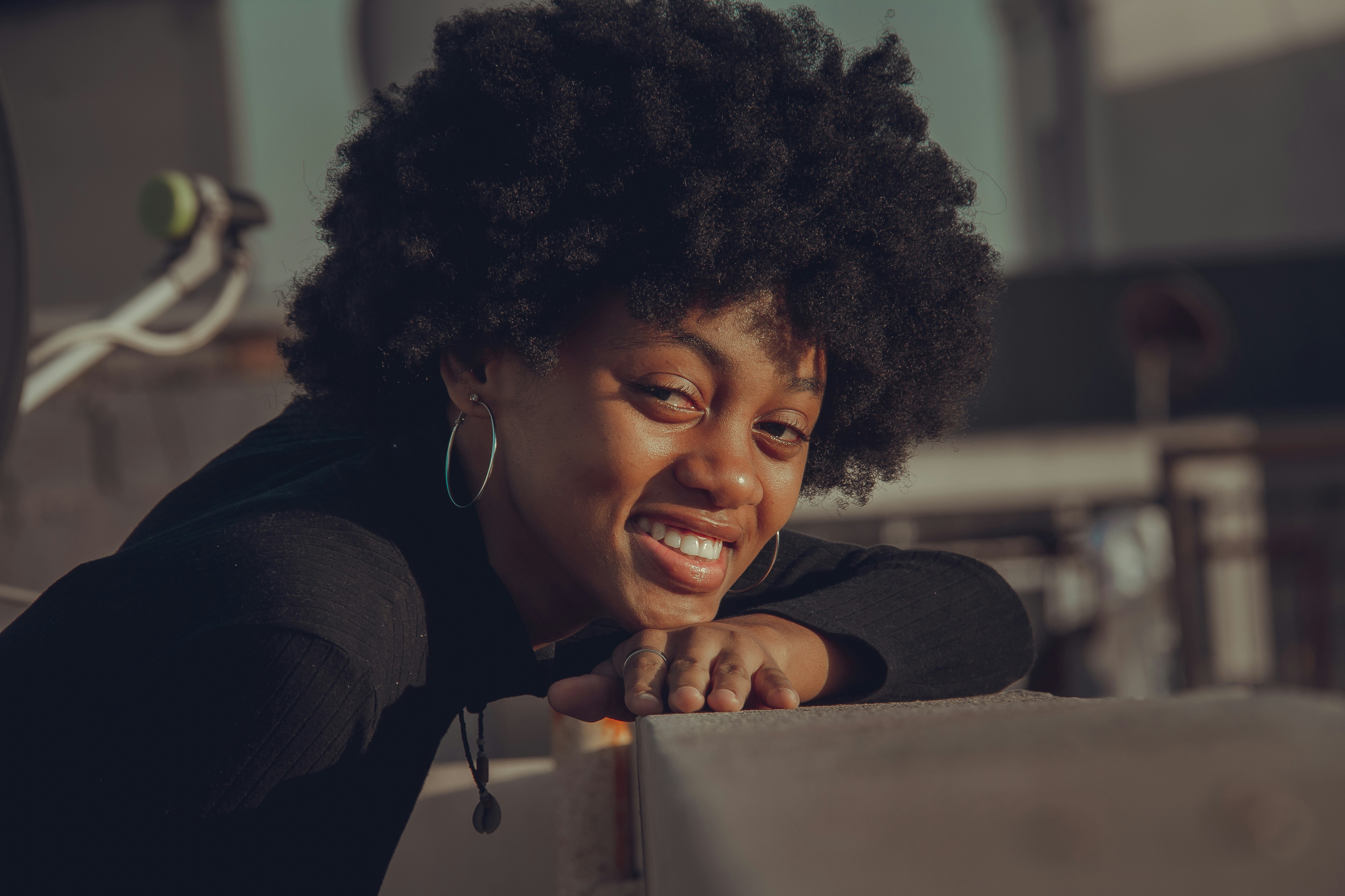 A young woman with curly hair smiles warmly while leaning on a railing in an urban setting, illuminated by soft natural light.