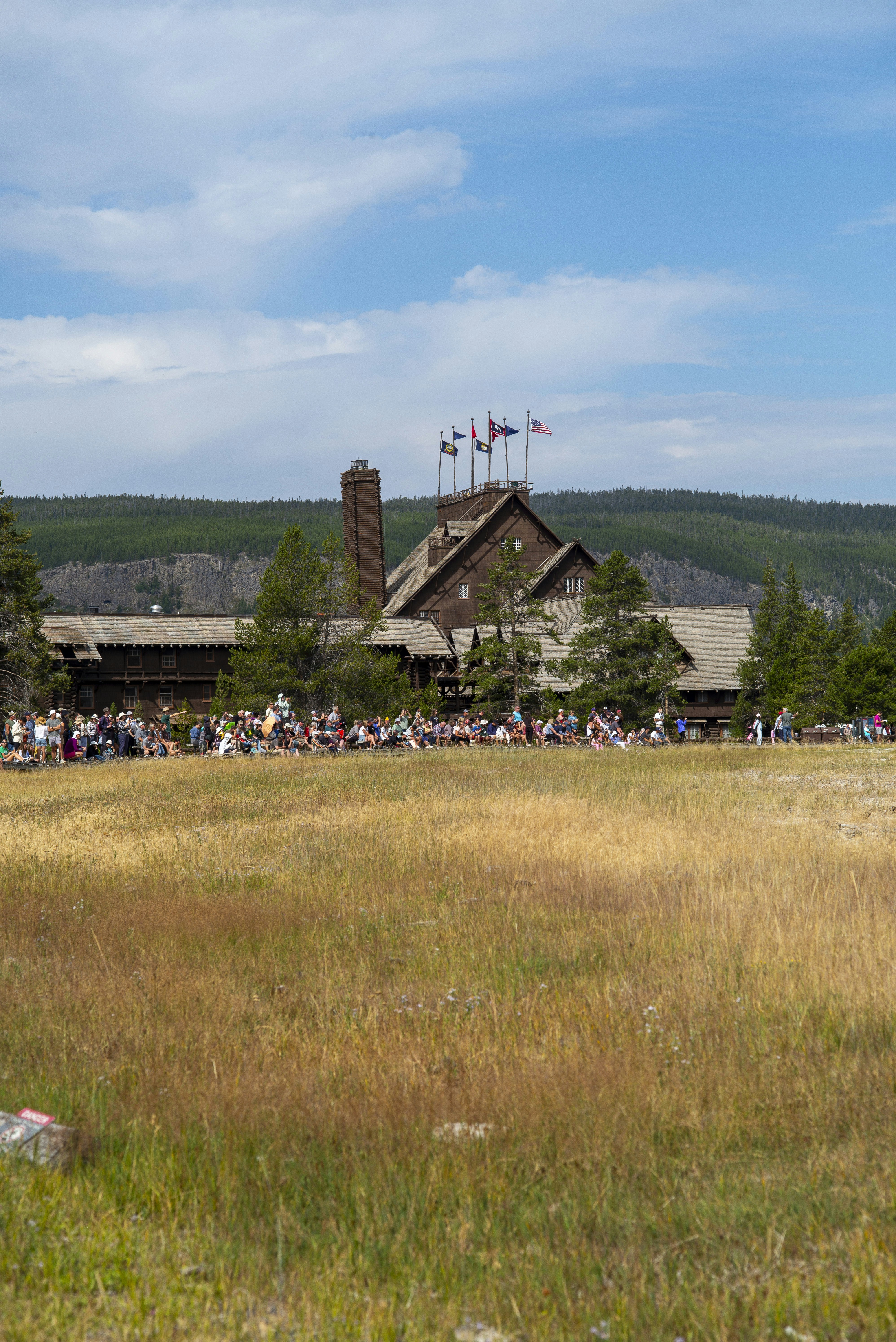 Crowd of visitors gathered in front of a historic lodge surrounded by lush greenery and distant mountains.