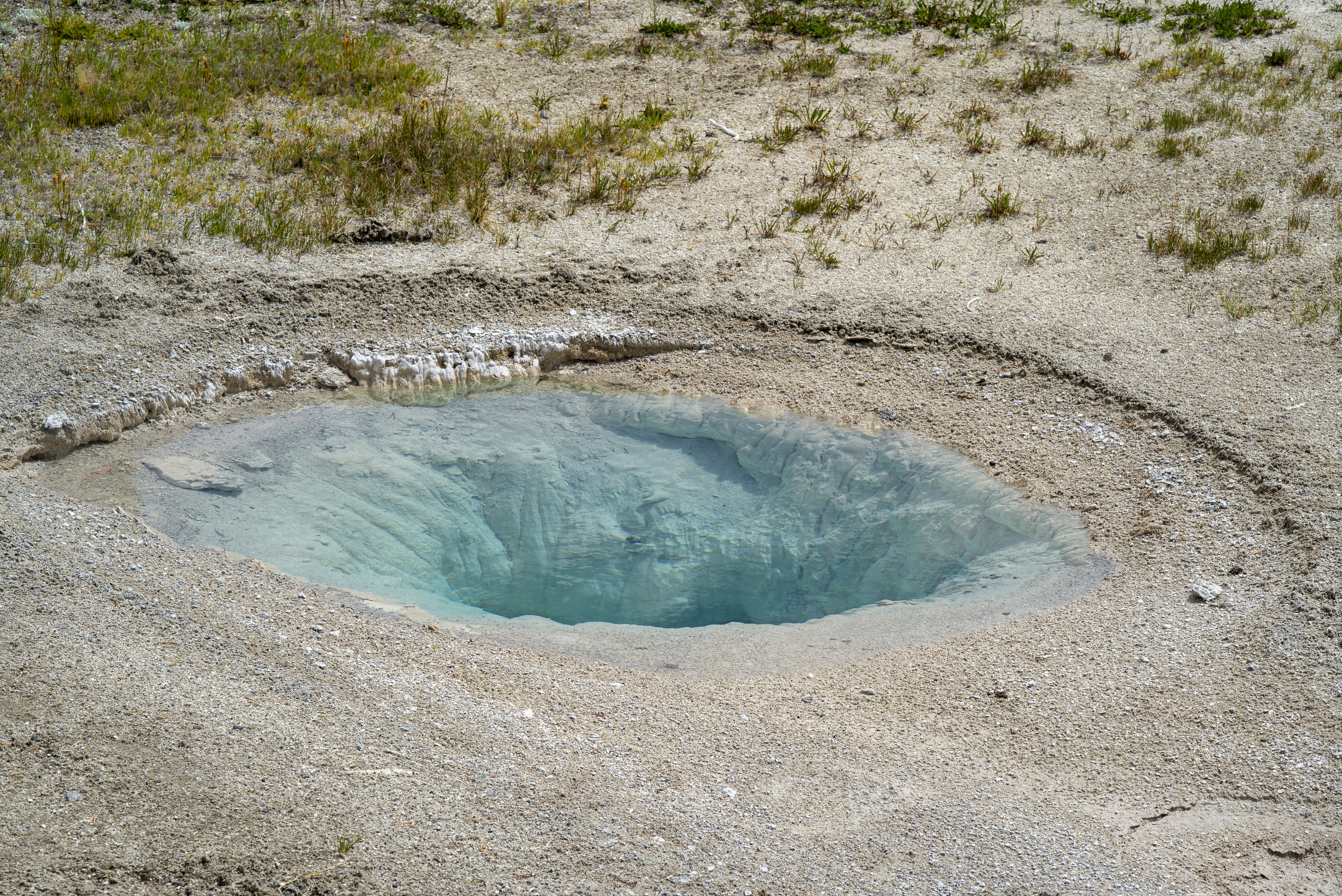 Crystal-clear geothermal pool surrounded by sandy terrain and sparse vegetation. The unique geological formation invites exploration.
