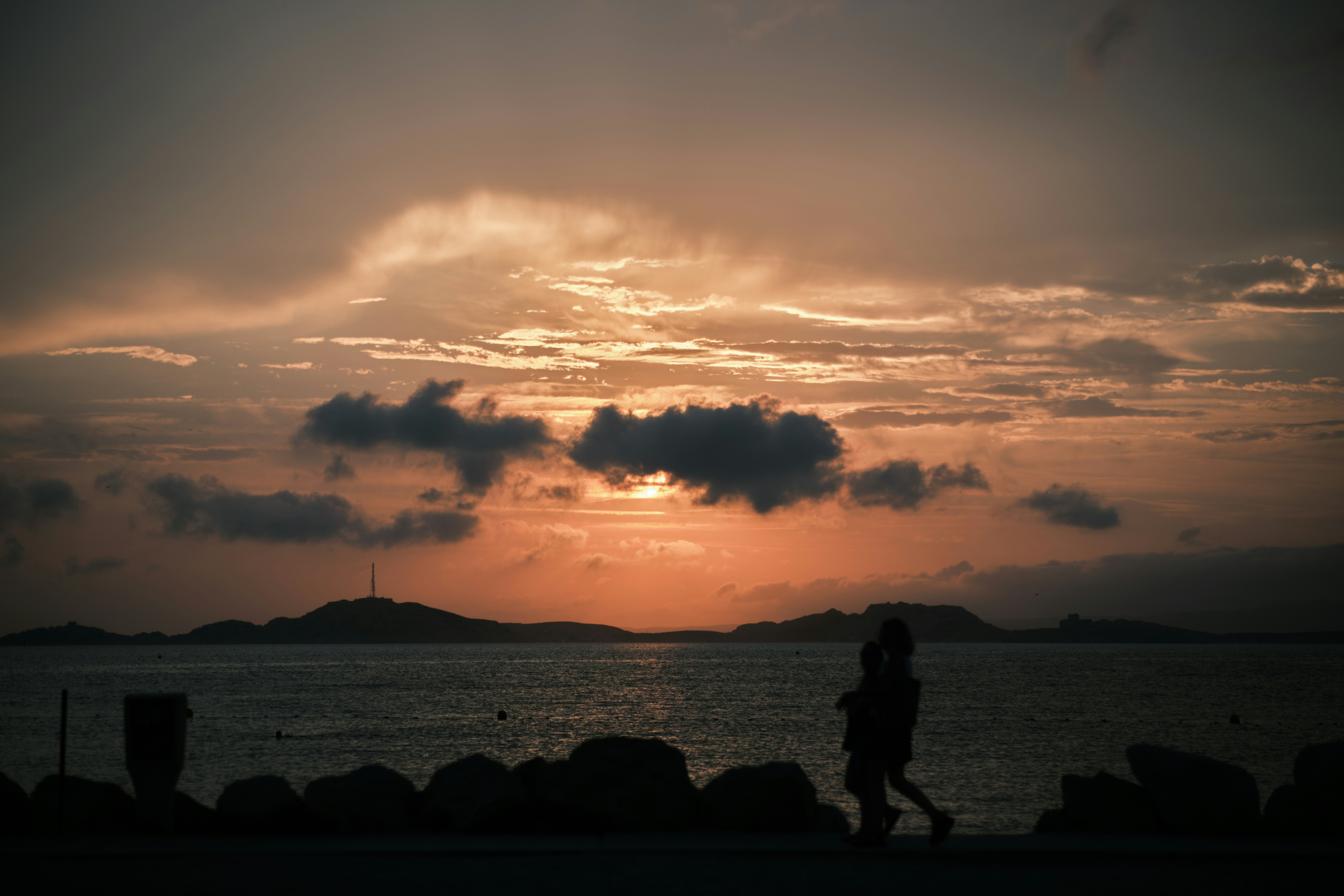 Sunset with walking silhouettes by the sea. Warm sunset over the sea with the silhouette of a couple walking along the waterfront, distant islands on the horizon, and dramatic clouds. | Two people walking along the coast at sunset.