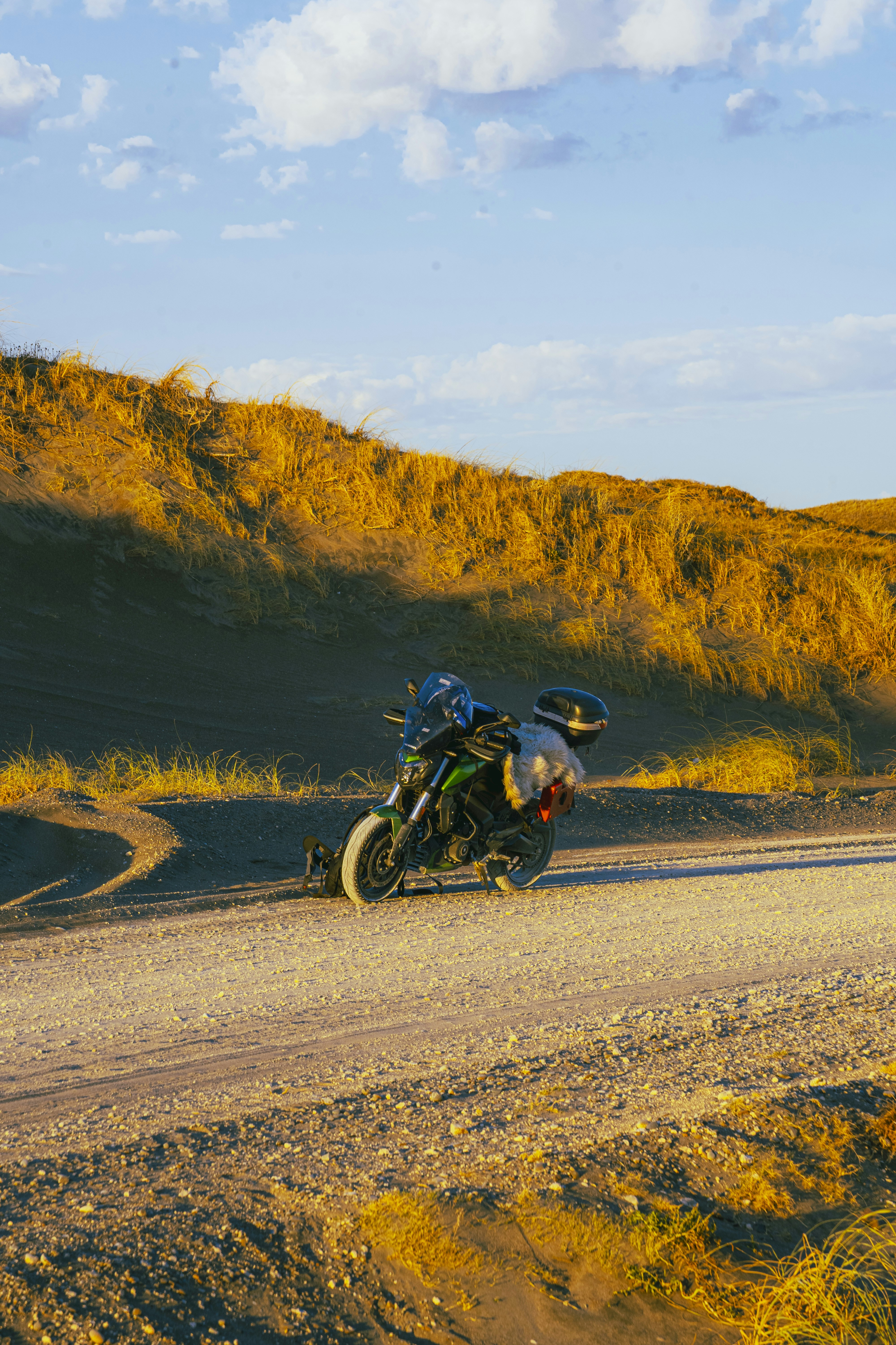 Motorcycle parked on a winding gravel path with golden grass and hills under a blue sky with fluffy clouds.