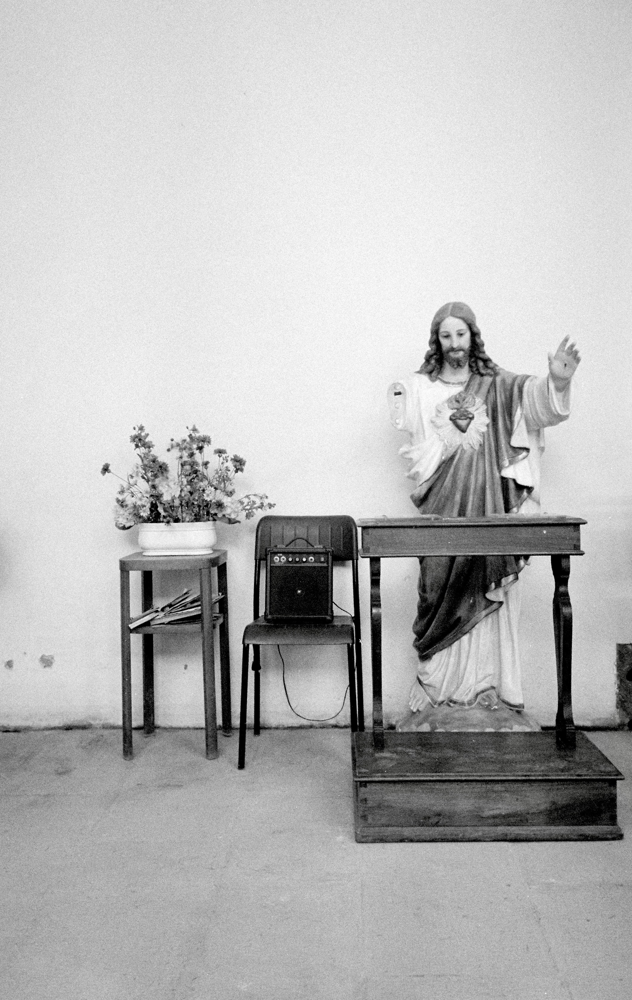 A serene interior scene featuring a statue of Jesus beside a small altar and a chair, complemented by a vase of flowers. The simplicity evokes a sense of tranquility.