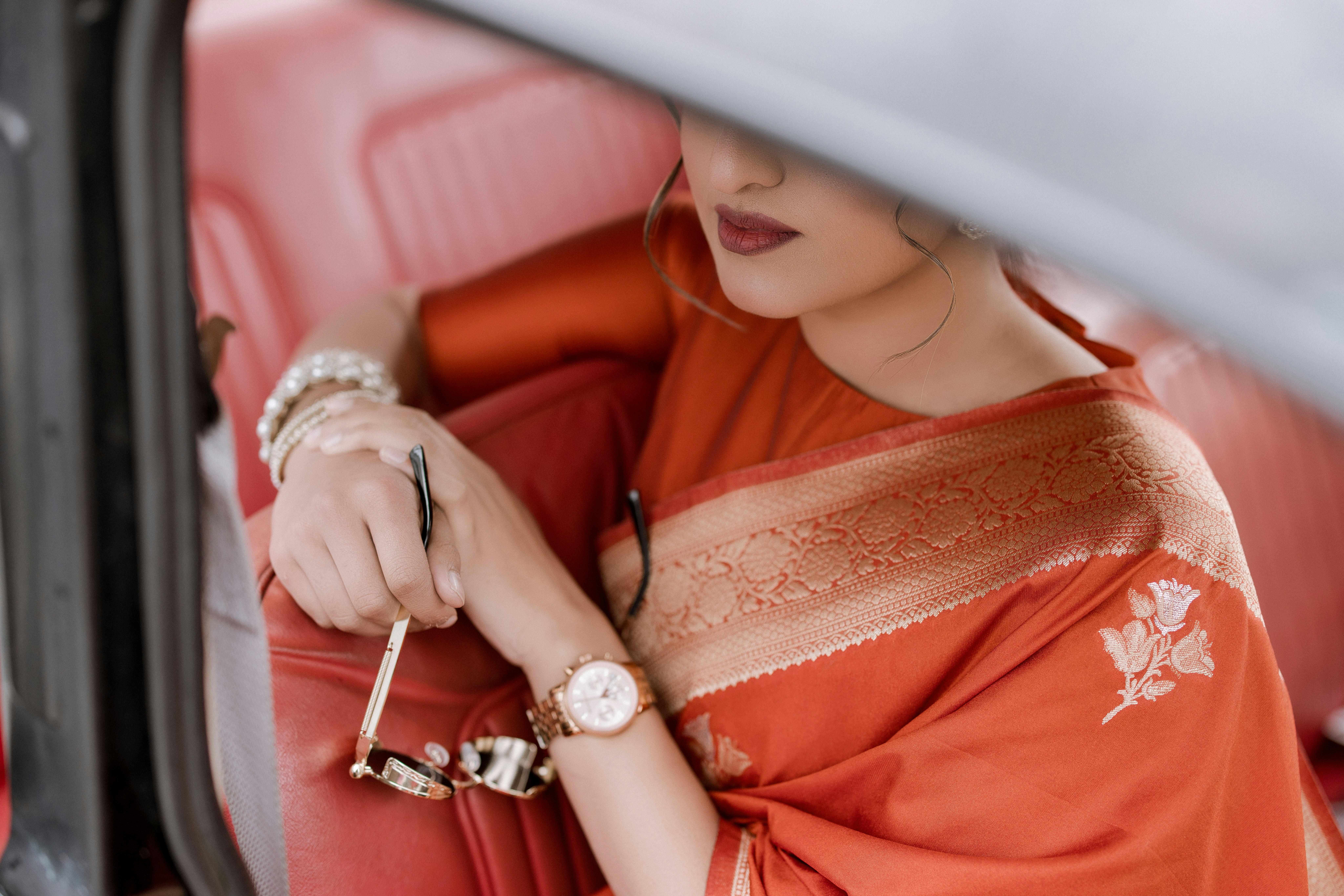 A woman in a vibrant orange saree, seated in a car, delicately holds a pen and watch while exuding grace and poise.