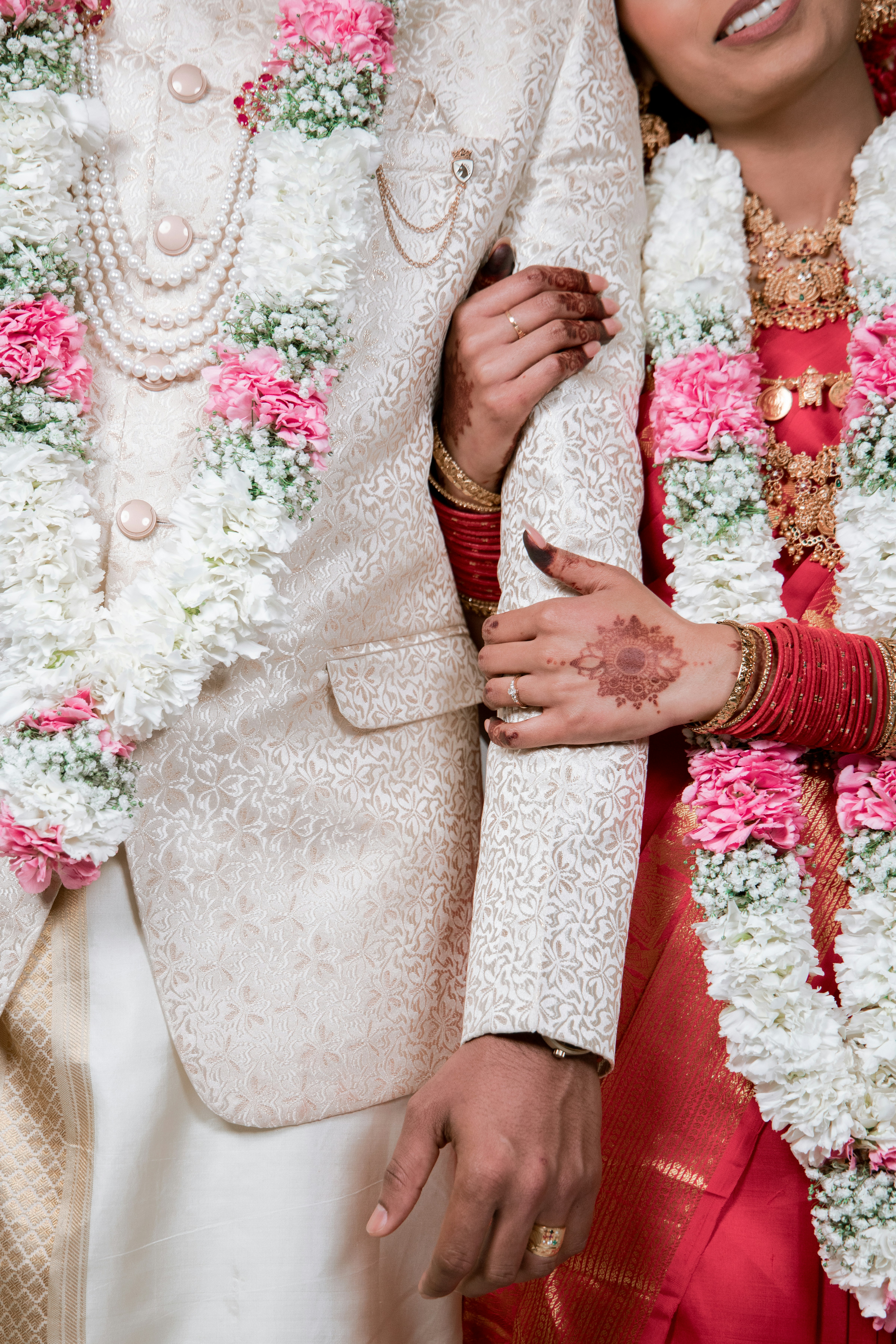 Close-up of a couple adorned in traditional attire, showcasing intricate henna and floral garlands. Their hands intertwined symbolize unity and commitment.