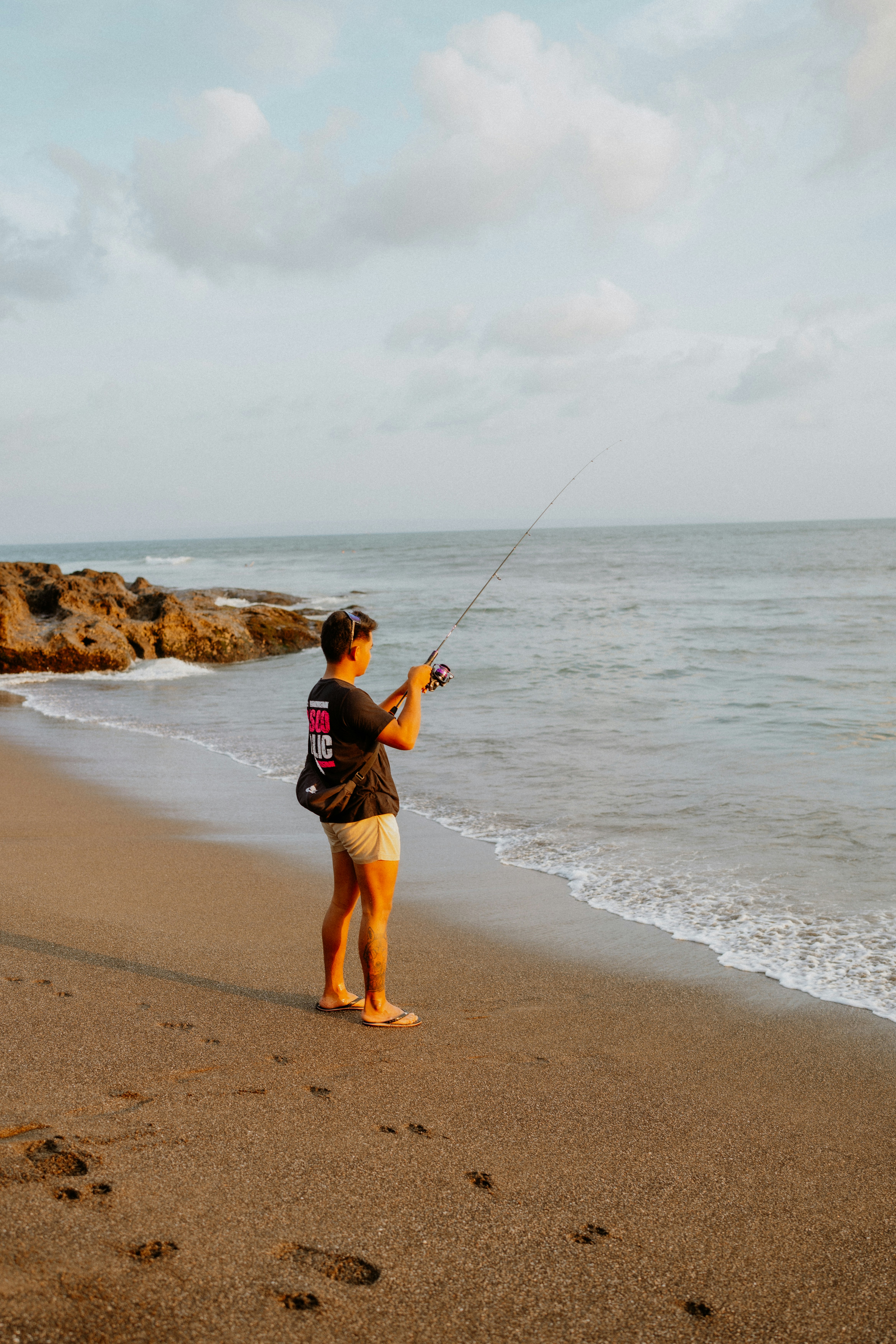 A man fishes along the sandy shore of Pererenan Beach, Bali during the early evening light. A calm scene of oceanfront leisure. | Man fishing on a sandy beach by the ocean