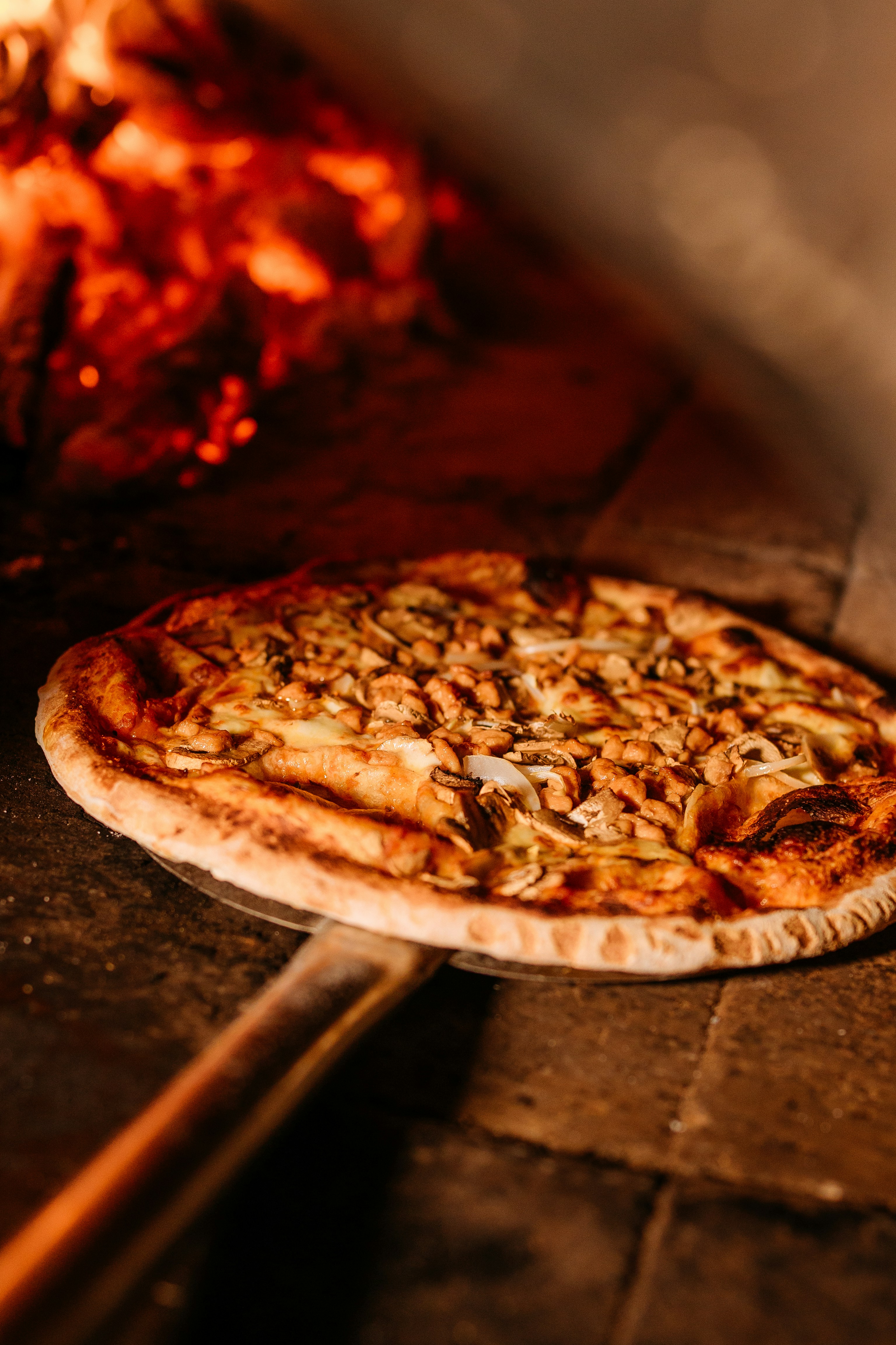A freshly topped pizza baking inside a traditional wood-fired oven in Gianyar, Bali, with flames creating the perfect crispy crust. | Pizza being removed from a wood-fired oven