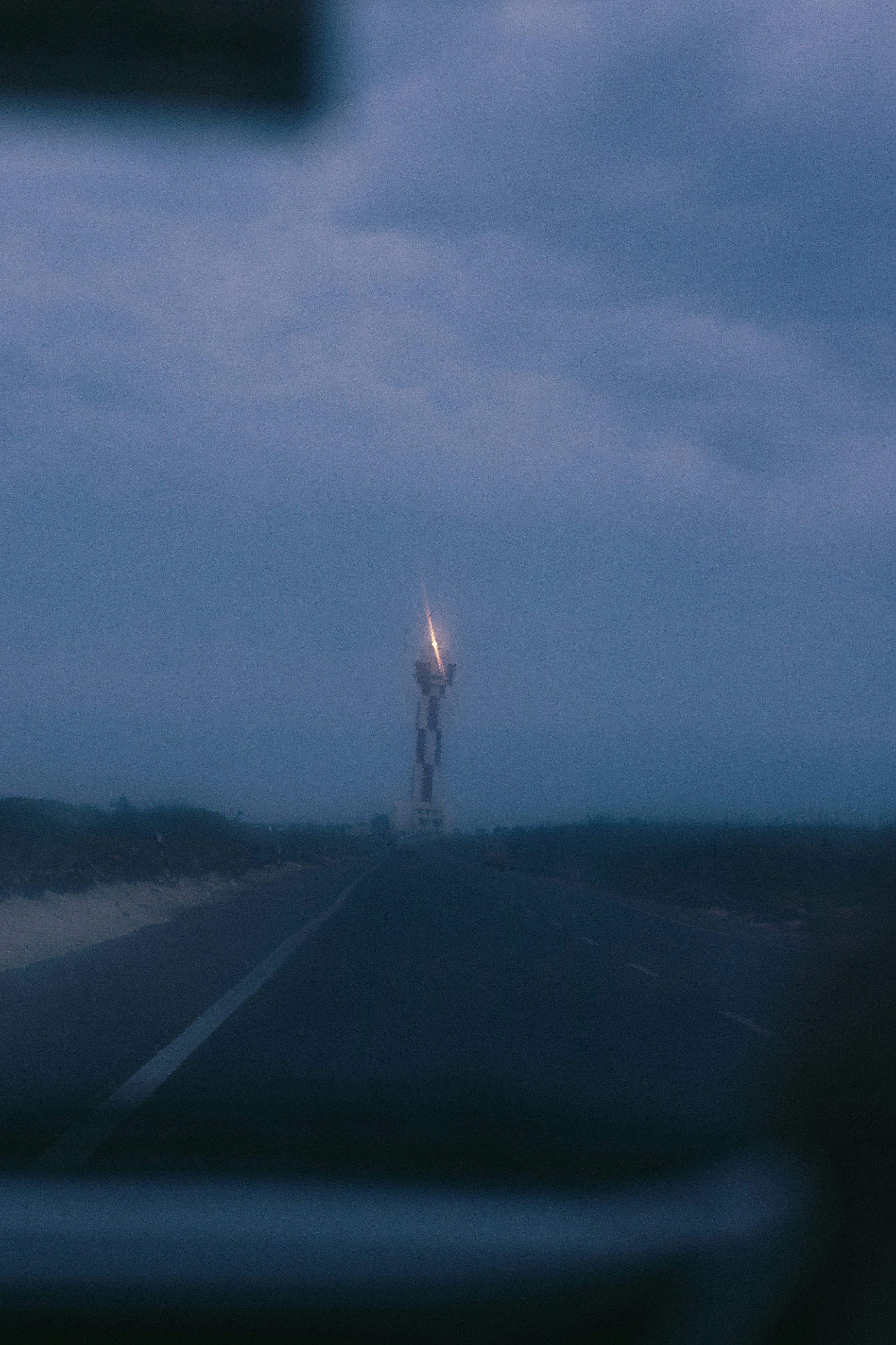 A distant lighthouse stands tall against a moody sky, illuminated by a beam of light. The empty road leads the viewer's gaze toward the beacon.