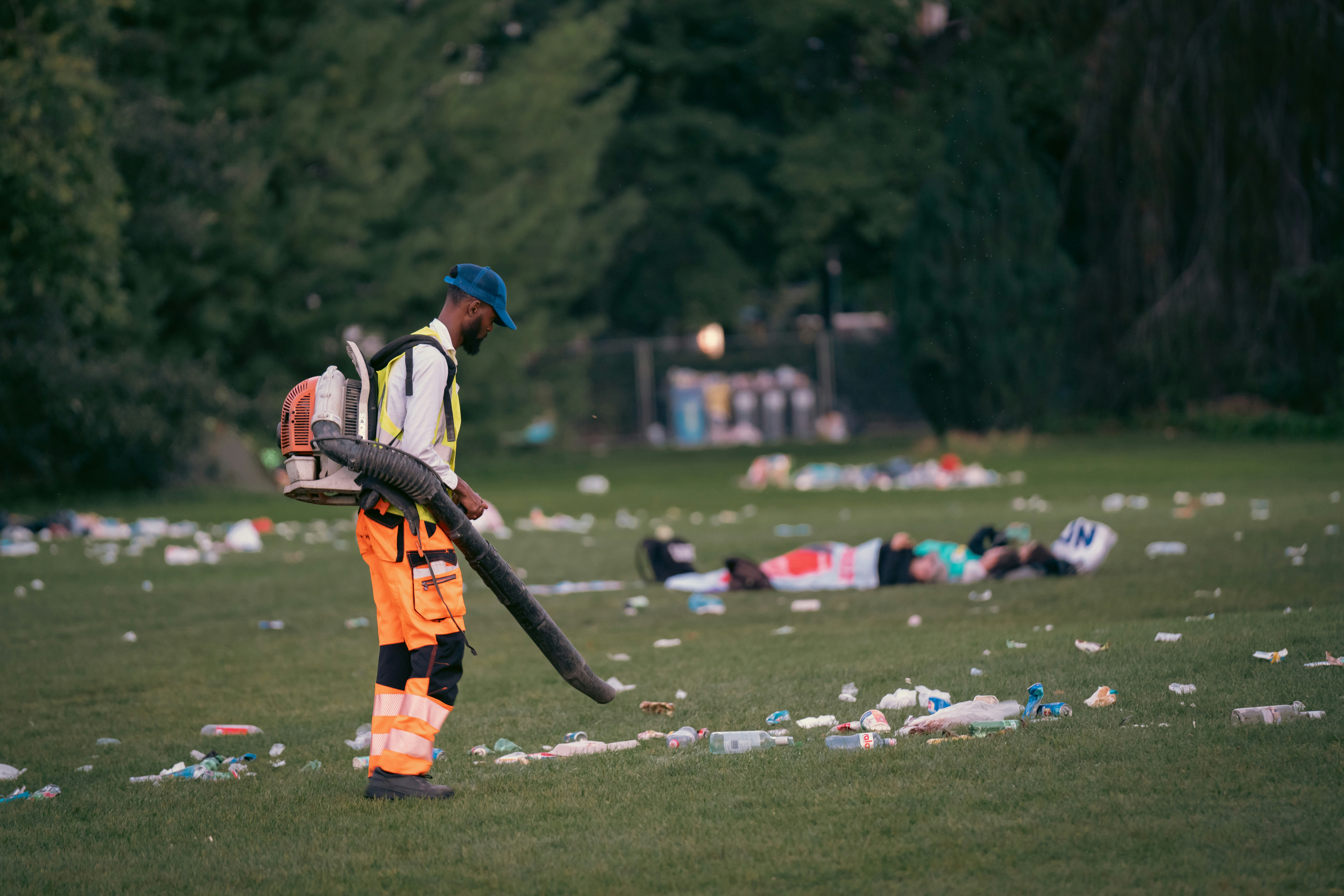 A groundskeeper using a leaf blower amidst scattered litter in a park, highlighting the aftermath of a busy event. The scene captures the contrast between nature and human impact.