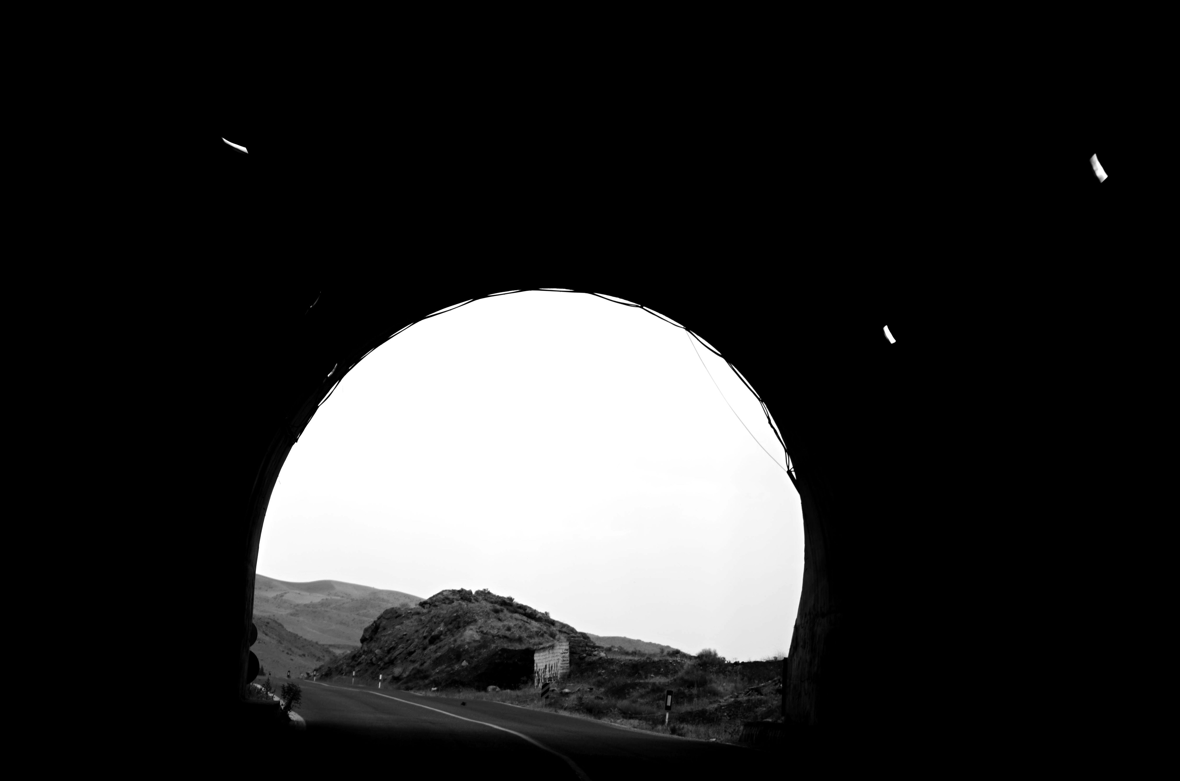 Black and white view through a tunnel, revealing a distant landscape beyond the circular opening. The contrast highlights the tunnel's structure and the scenery ahead.