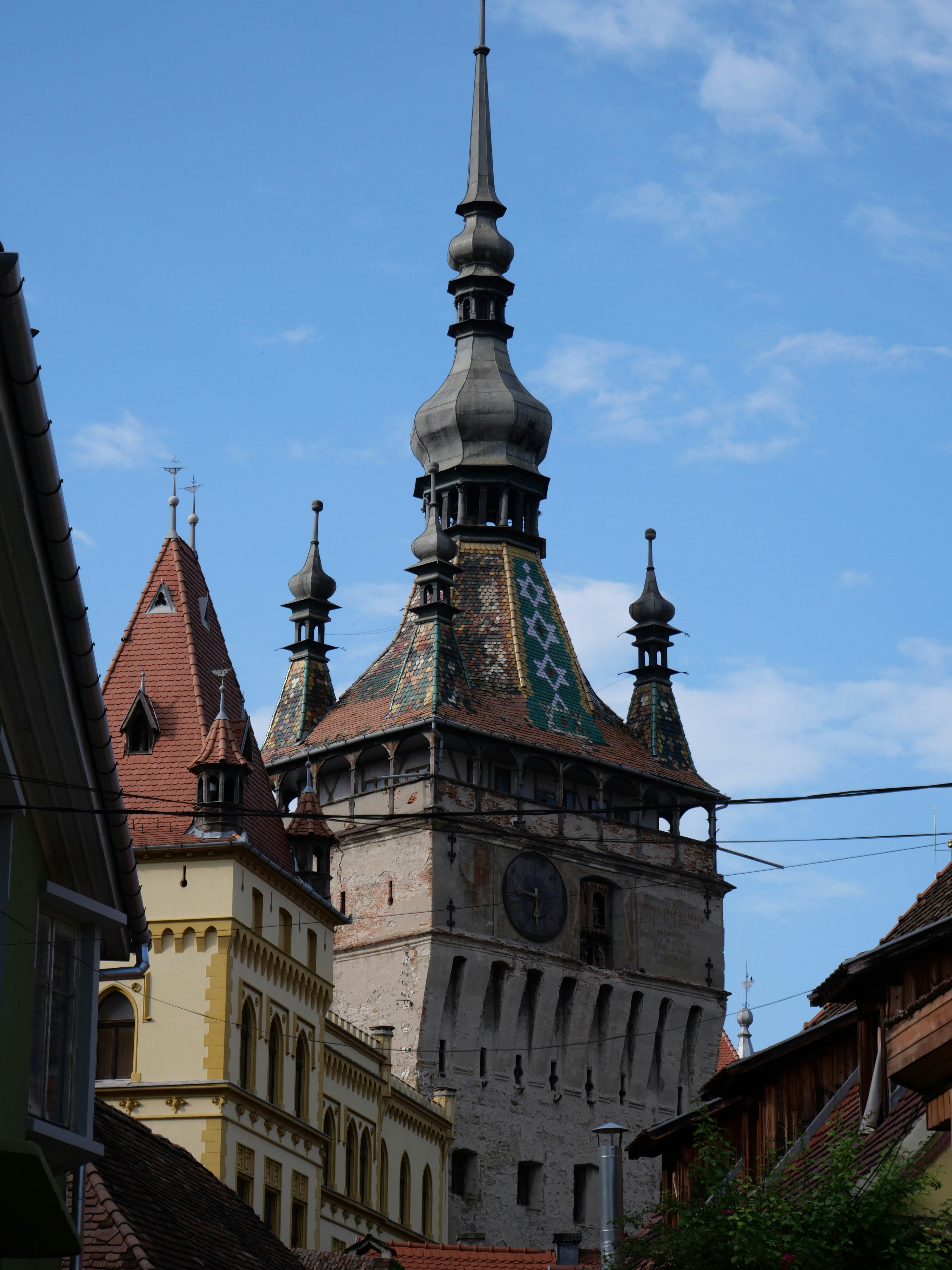 A close-up of the iconic Clock Tower (Turnul cu Ceas) in Sighișoara, Romania. This stunning piece of medieval architecture is the city's main landmark, with its colorful tiled roof and intricate details standing out against a bright blue sky. | Historic clock tower with surrounding buildings under blue sky