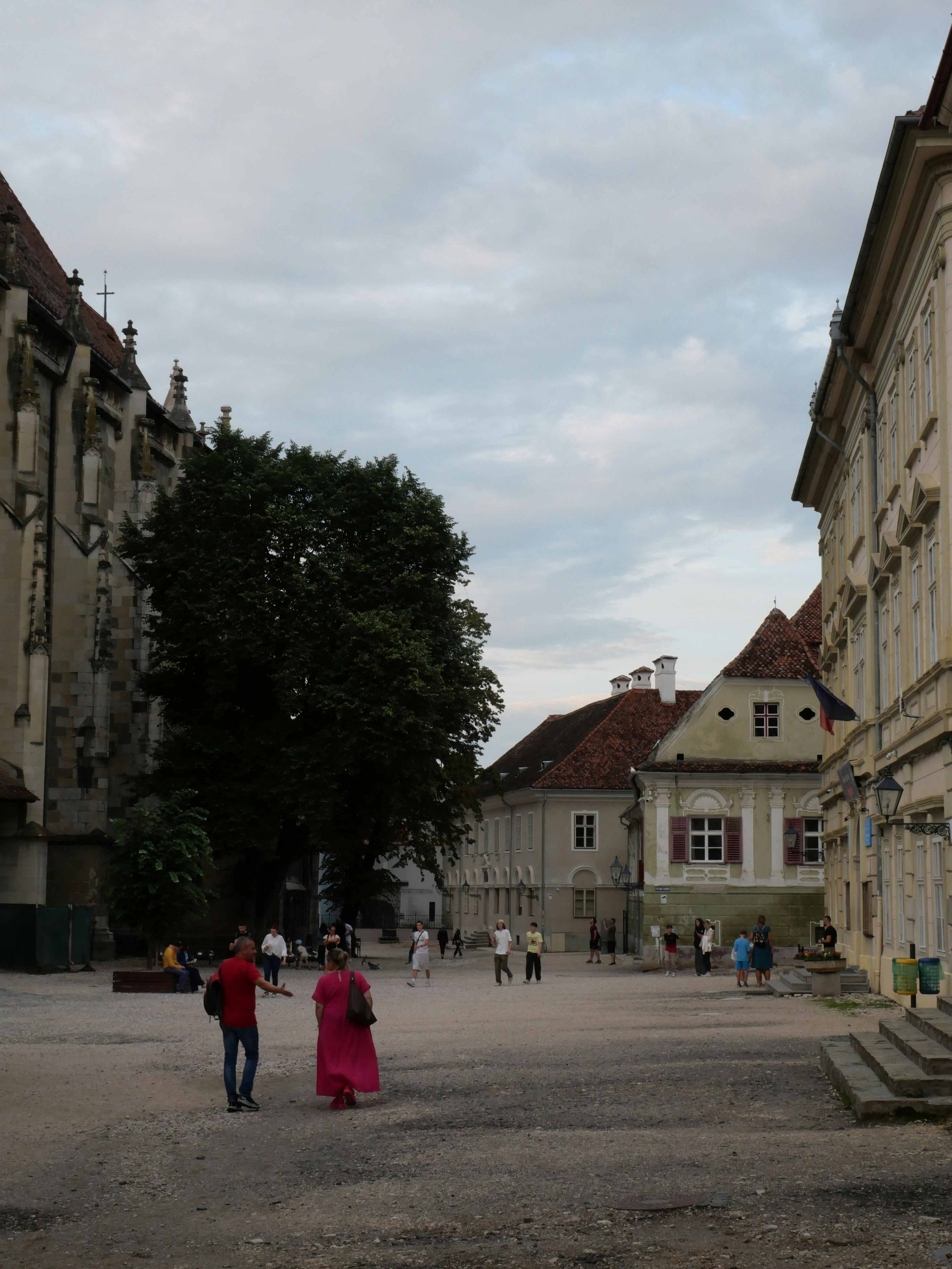 A pedestrian square in Brașov, Romania, bustling with activity. The photo captures people walking past historic buildings with traditional architecture, reflecting the lively atmosphere of the city's old town. | People walking in an open square with buildings around.