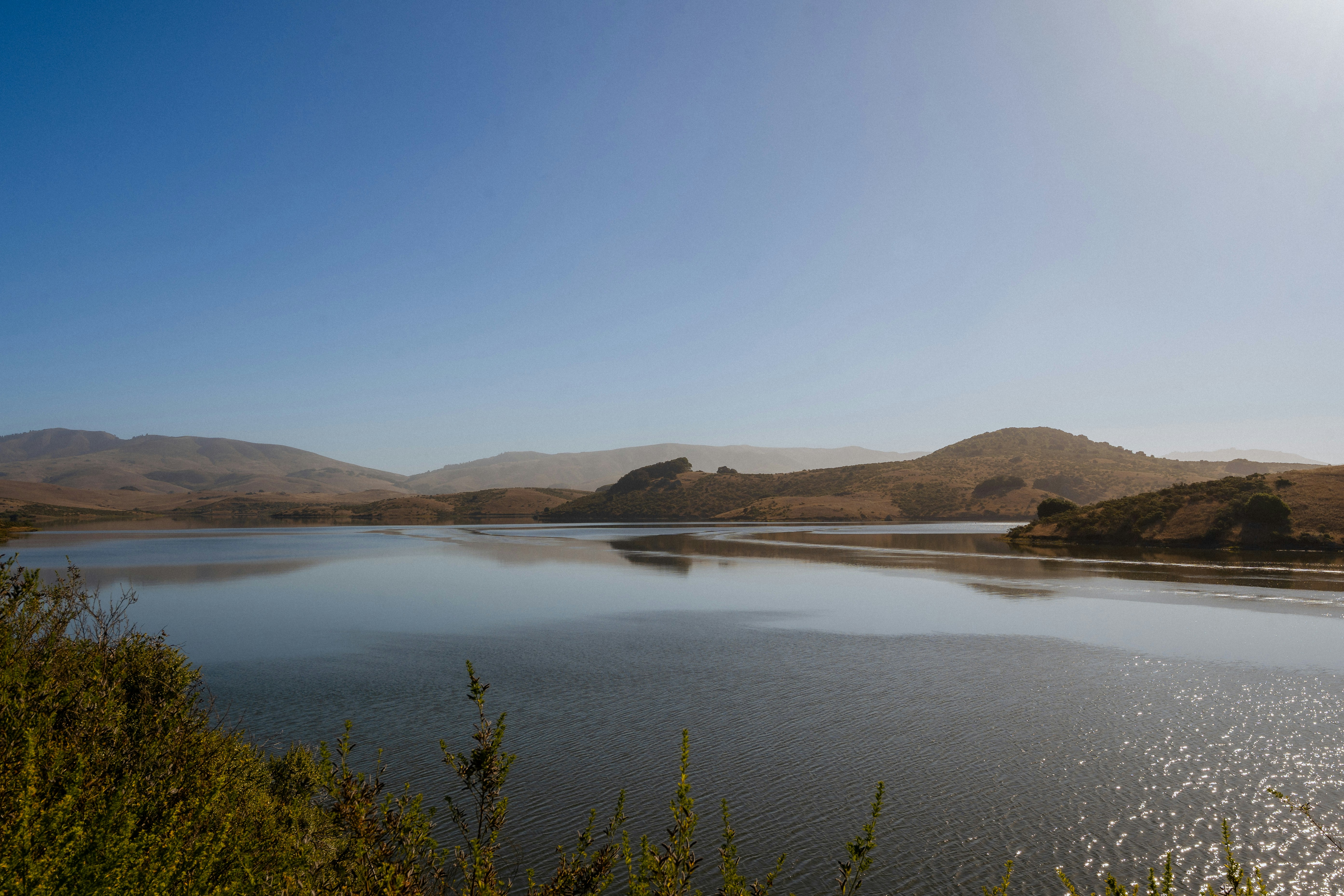 Calm waters reflect the rolling hills under a clear blue sky — a peaceful lakeside scene in California’s countryside. | Calm lake reflecting hills under a clear sky