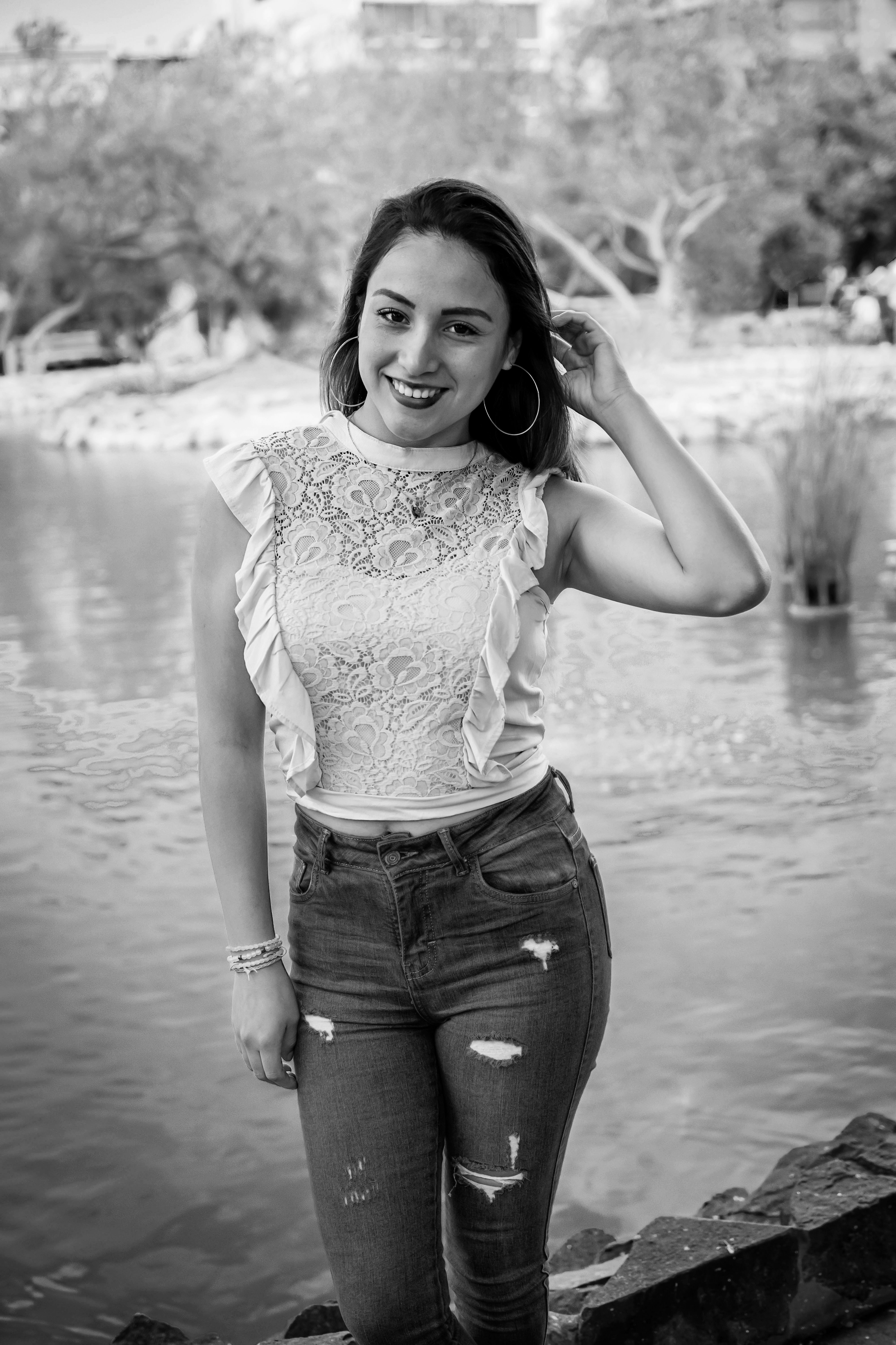 Young woman with a playful smile poses by a tranquil water body, showcasing her stylish outfit. The monochrome setting enhances the mood.