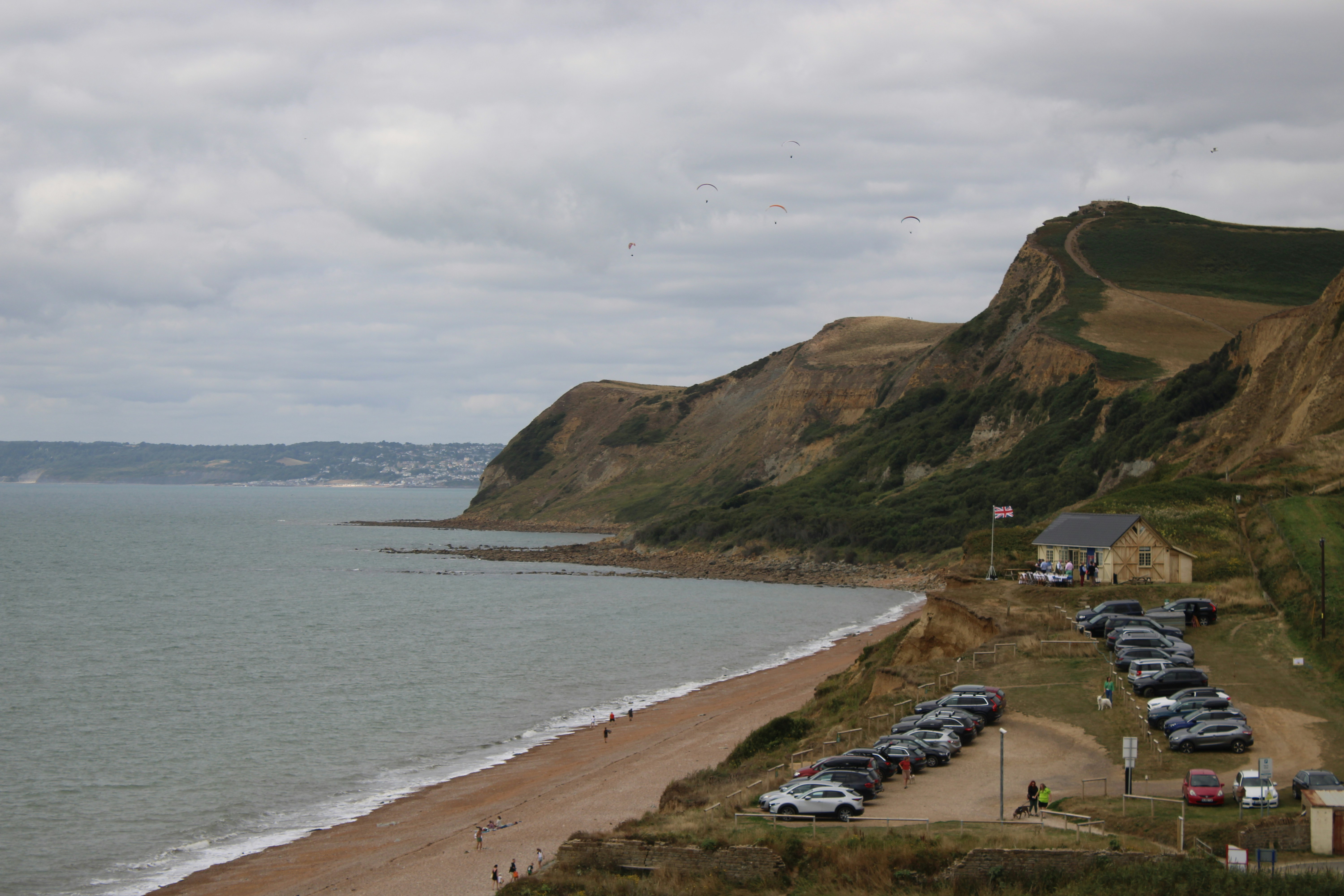 Waves gently lapping at the sandy beach, with a rugged cliff backdrop and a line of parked cars along the shore. A small building is visible, hinting at human presence in this natural landscape.