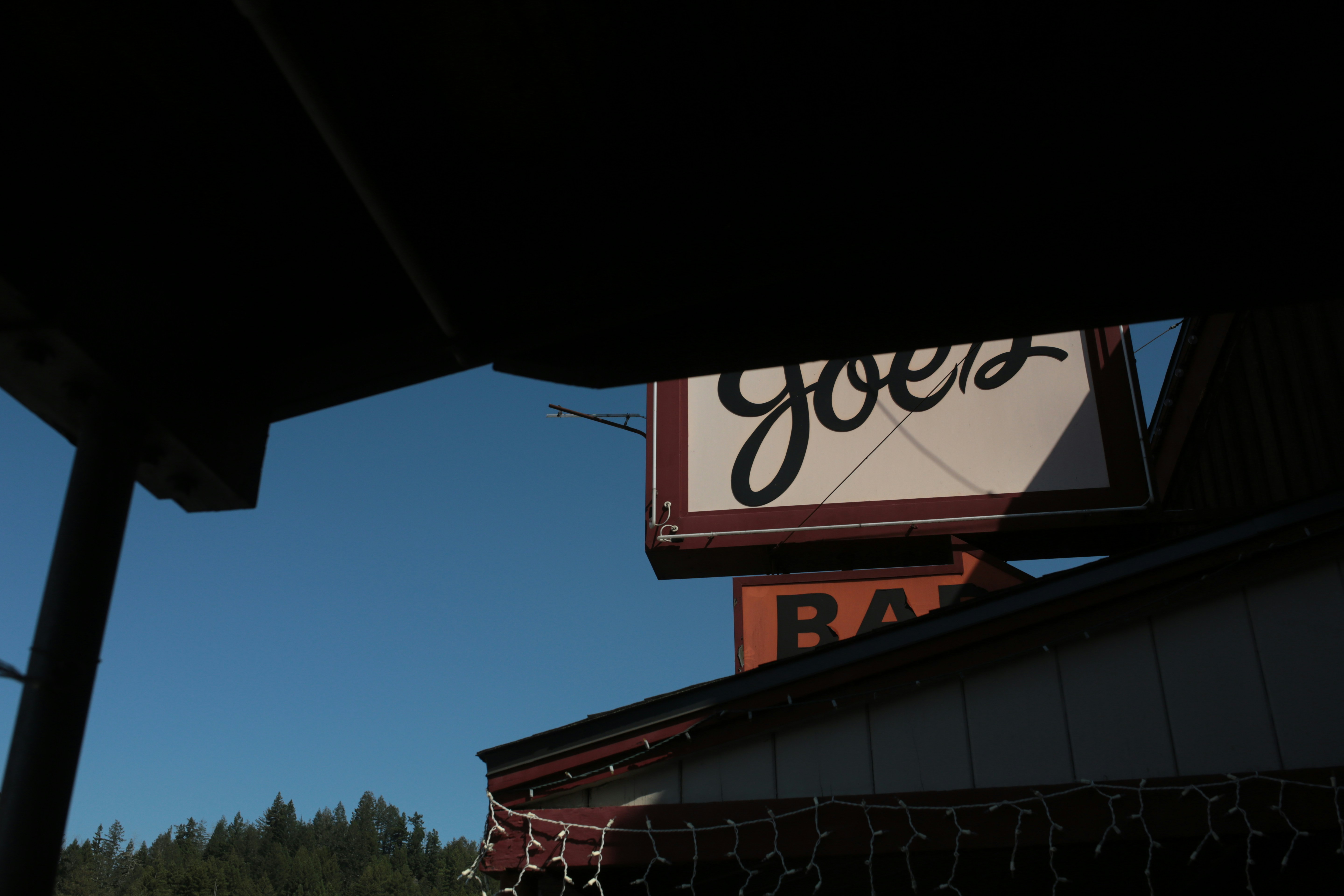 Signage of Joe's Bar partially obscured by a roof overhang, set against a clear blue sky with distant trees visible.