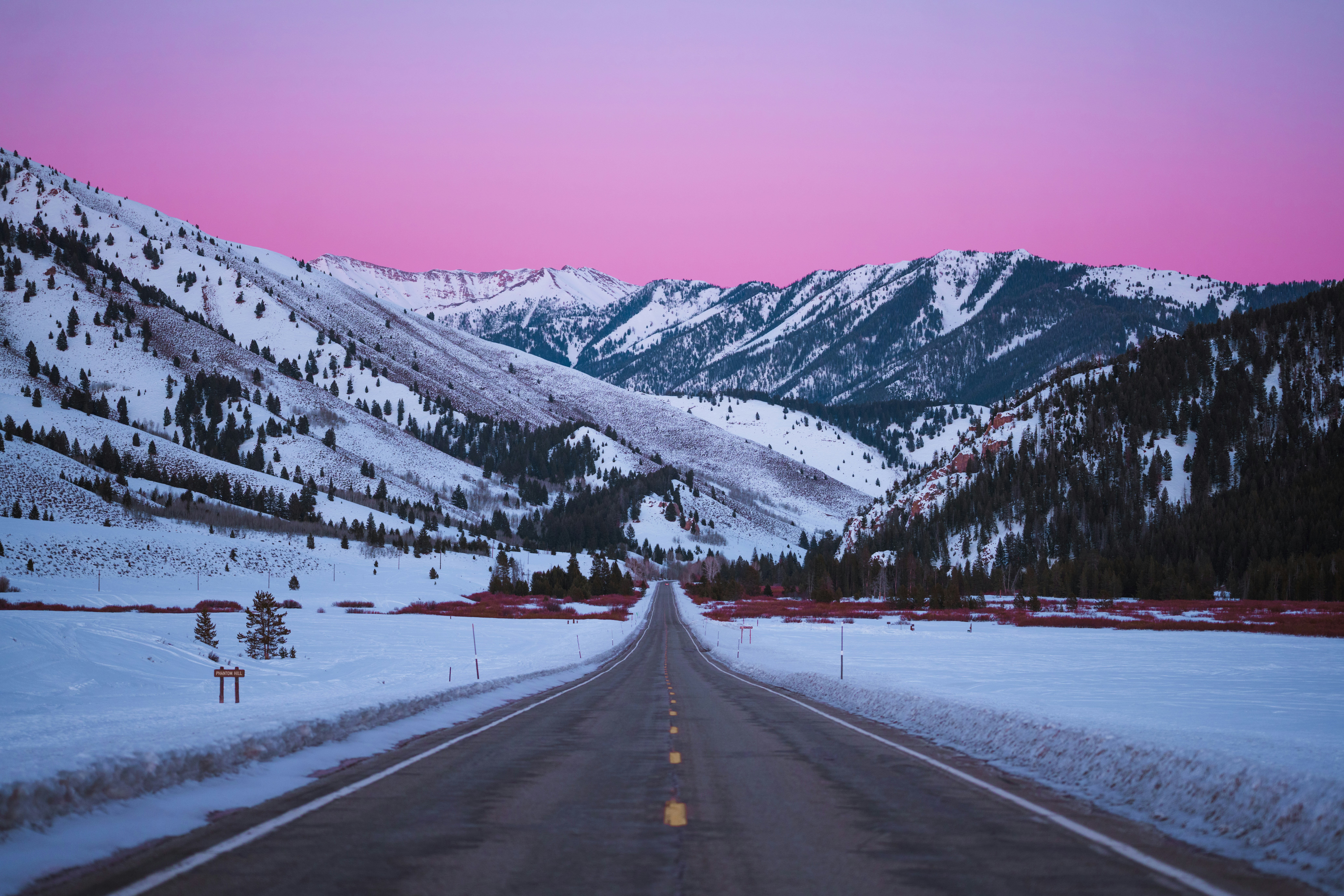 Scenic winter highway between Stanley and Ketchum, Idaho, framed by snow-covered Sawtooth Mountains and a stunning pink twilight sky. A picturesque mountain drive through the heart of Idaho’s wilderness. | Empty road through snow-covered mountains at dusk