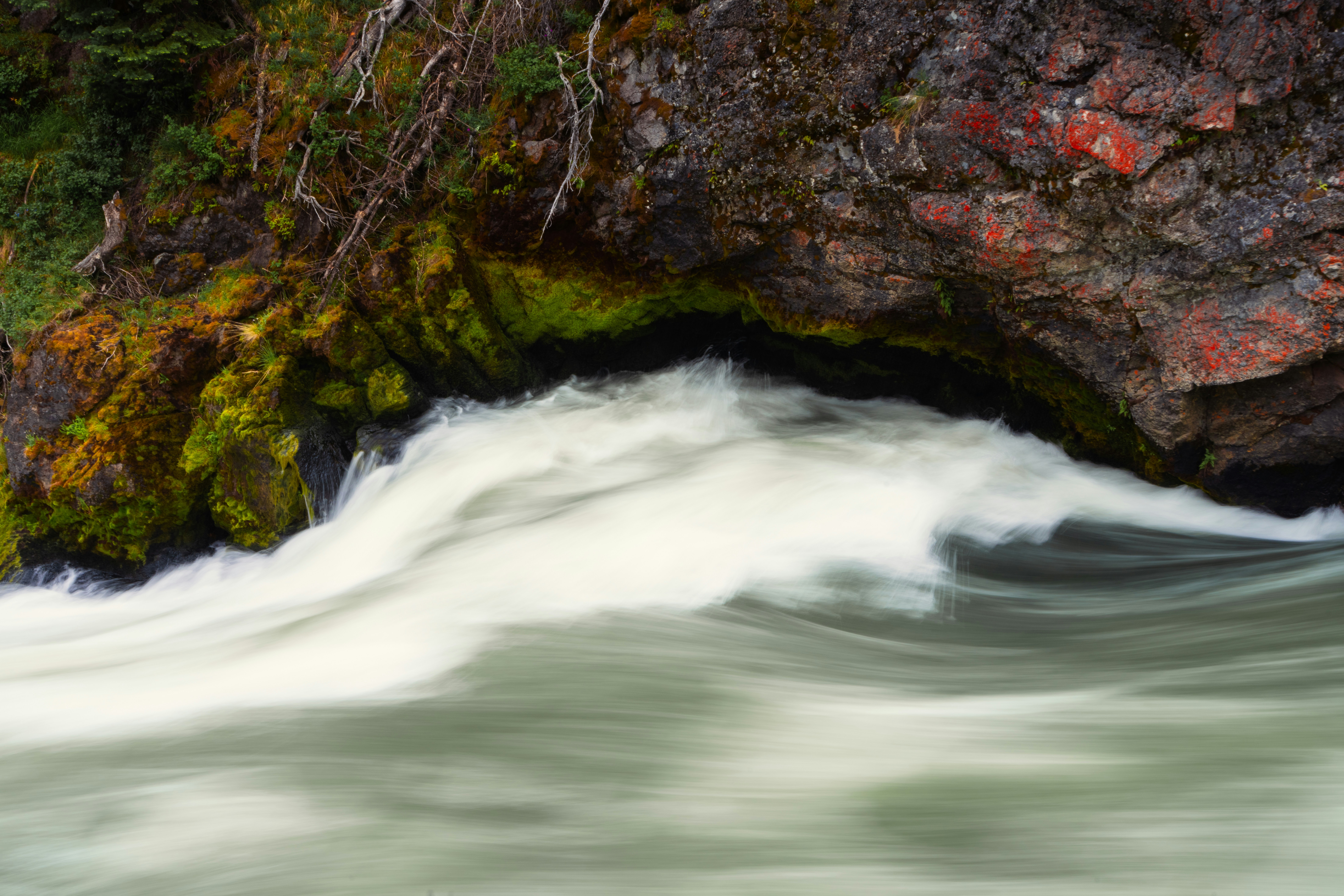 A powerful rush of whitewater flows past moss-covered rocks and rugged cliffs in Yellowstone National Park. The long-exposure effect captures the smooth motion of the water as it swirls and curves, contrasting with the vivid green moss and rusty red lichen on the surrounding stone. | White water rapids flow past mossy rocks